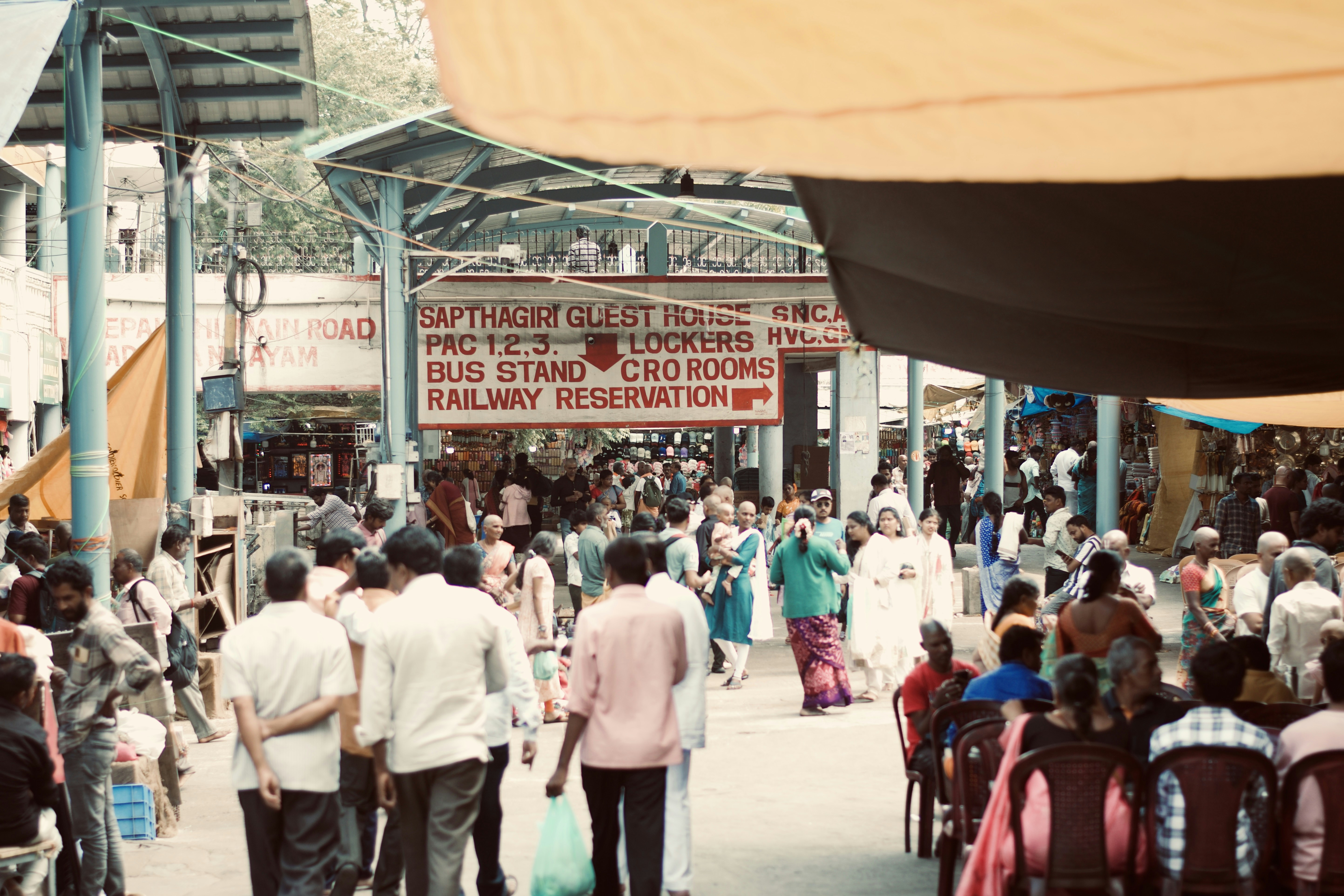 Crowded market area with people moving under a metal canopy, surrounded by signs for guest houses and railway reservation.