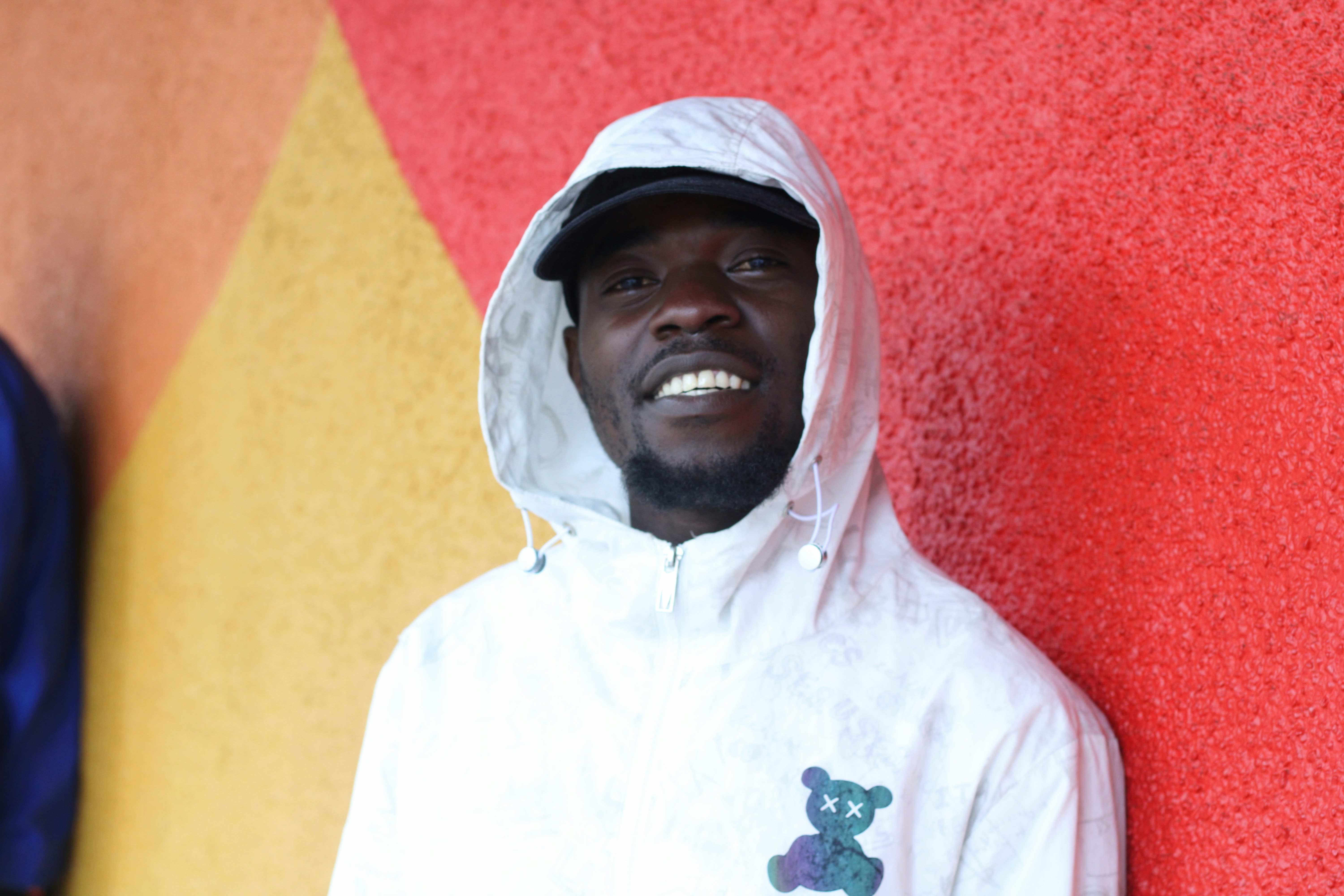 Person in a white hoodie with a playful bear patch stands against a colorful textured wall.