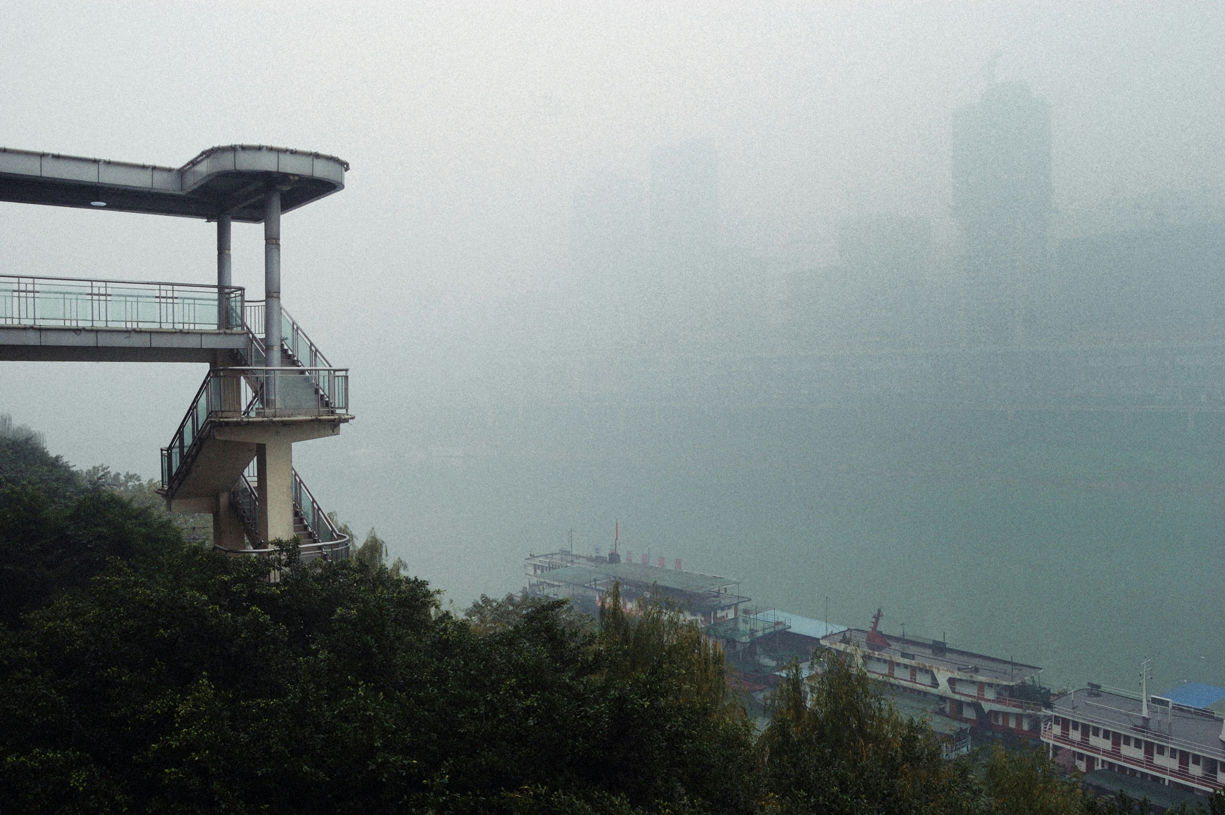 Foggy scene with elevated walkway and moored boats along a hazy riverbank.
