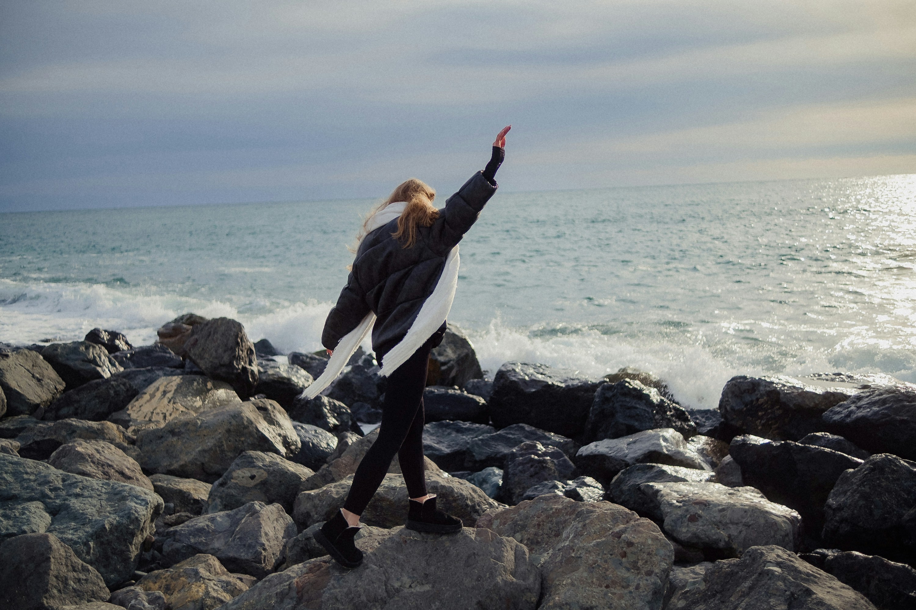 Person balancing on coastal rocks with ocean waves and a breezy sky.