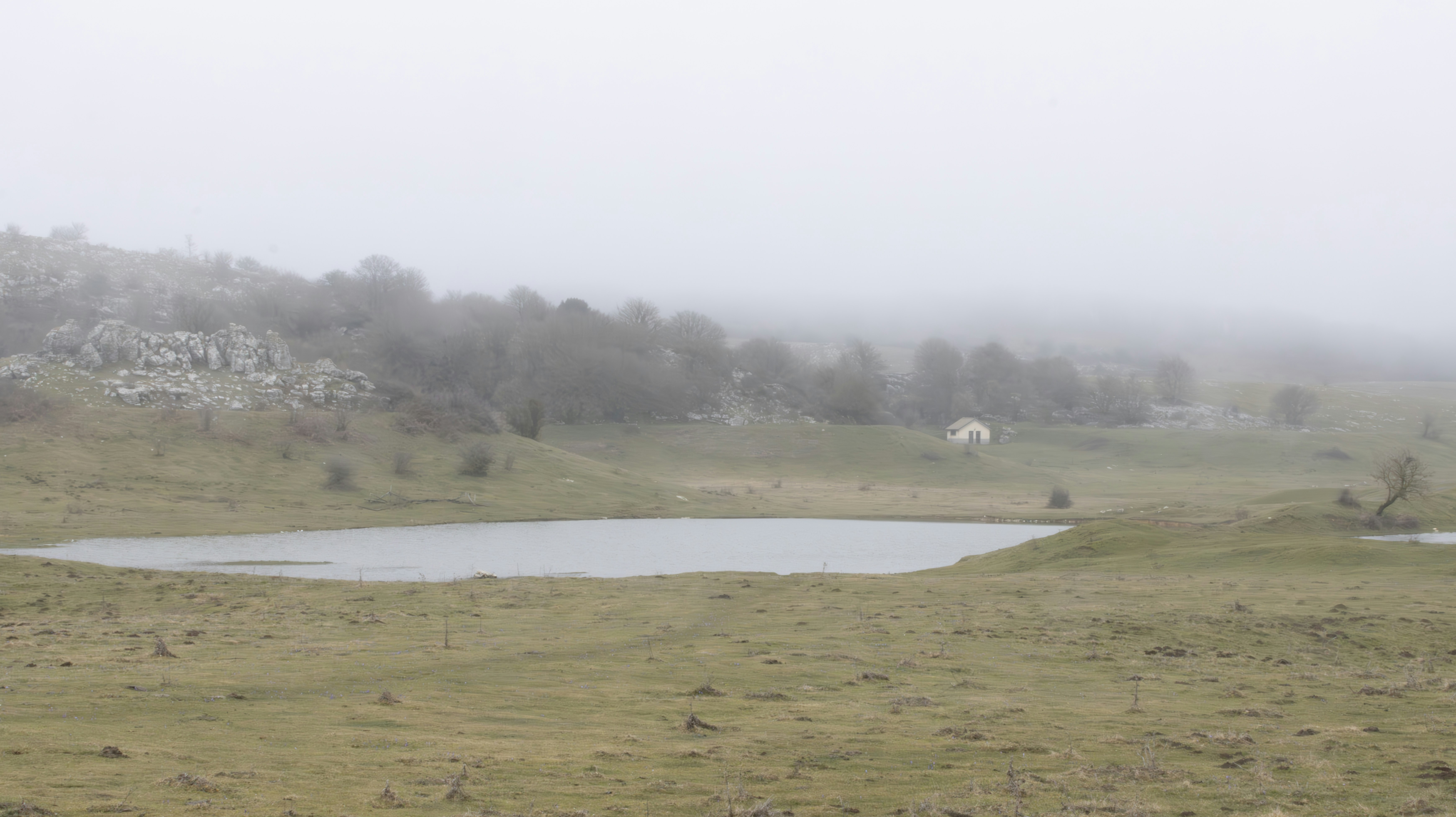Foggy landscape with a solitary house near a tranquil pond and distant hills.