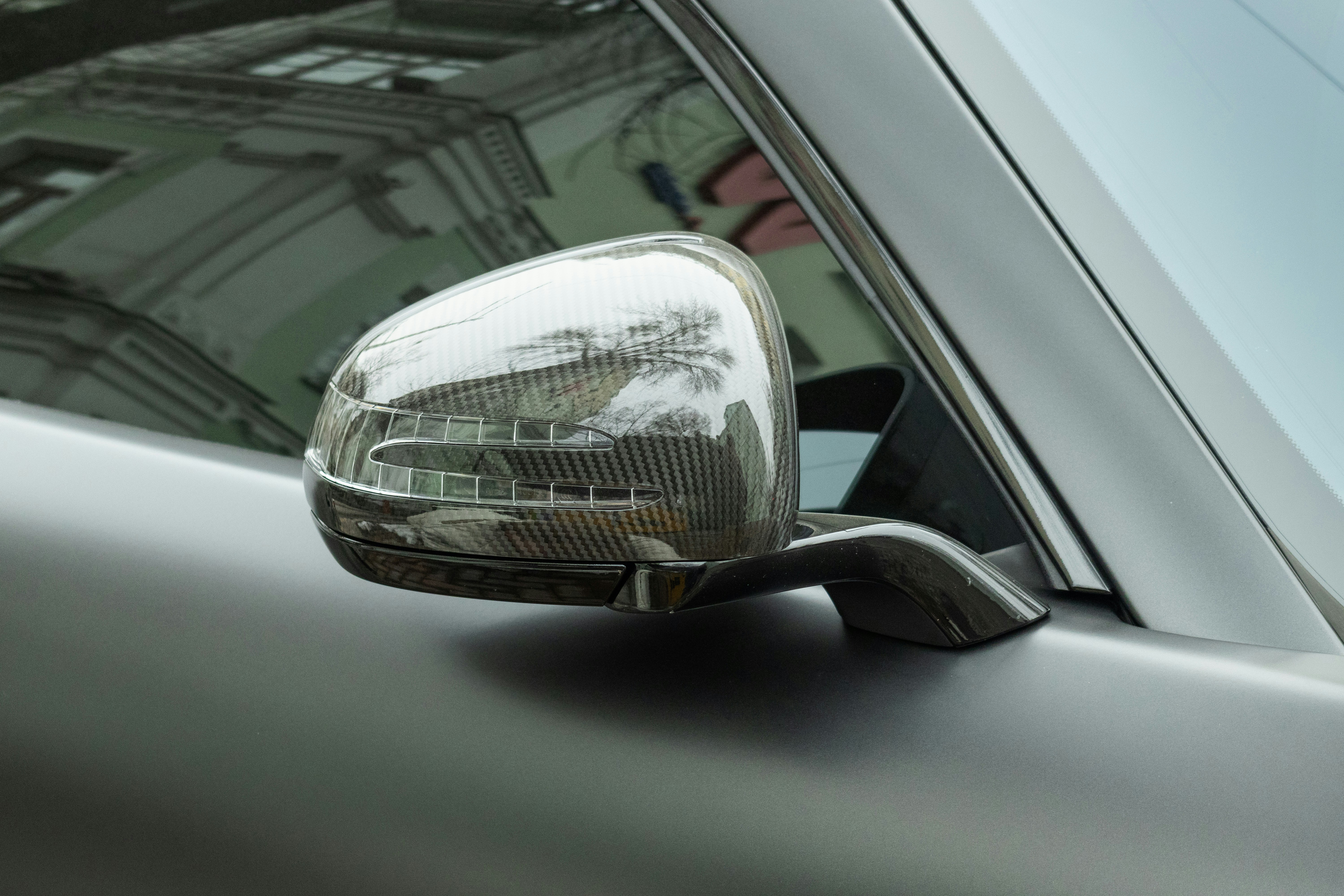 Close-up of a car's side mirror with a carbon fiber finish reflecting nearby architecture.