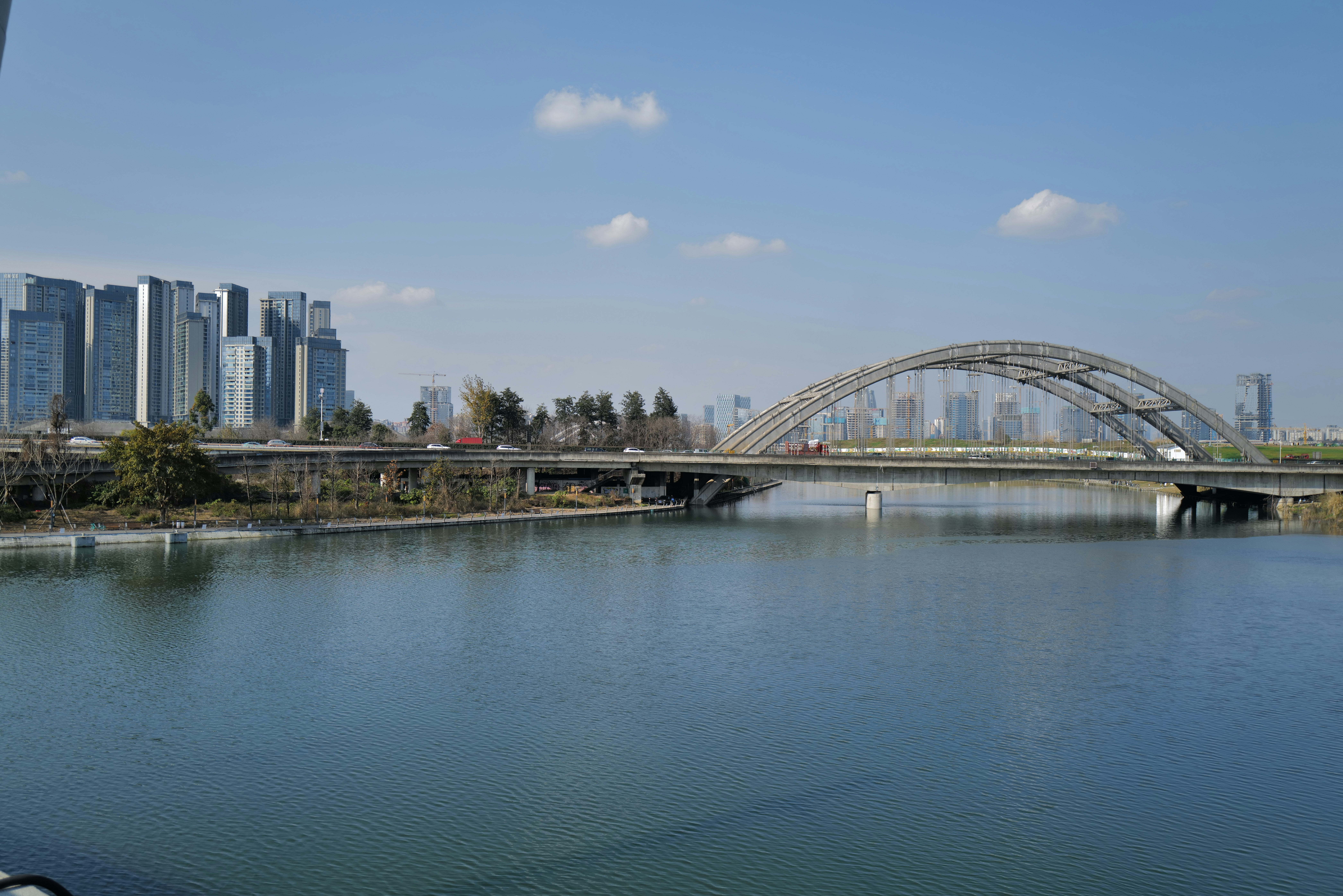 Modern bridge arches over a river with a backdrop of high-rise buildings under a clear blue sky.