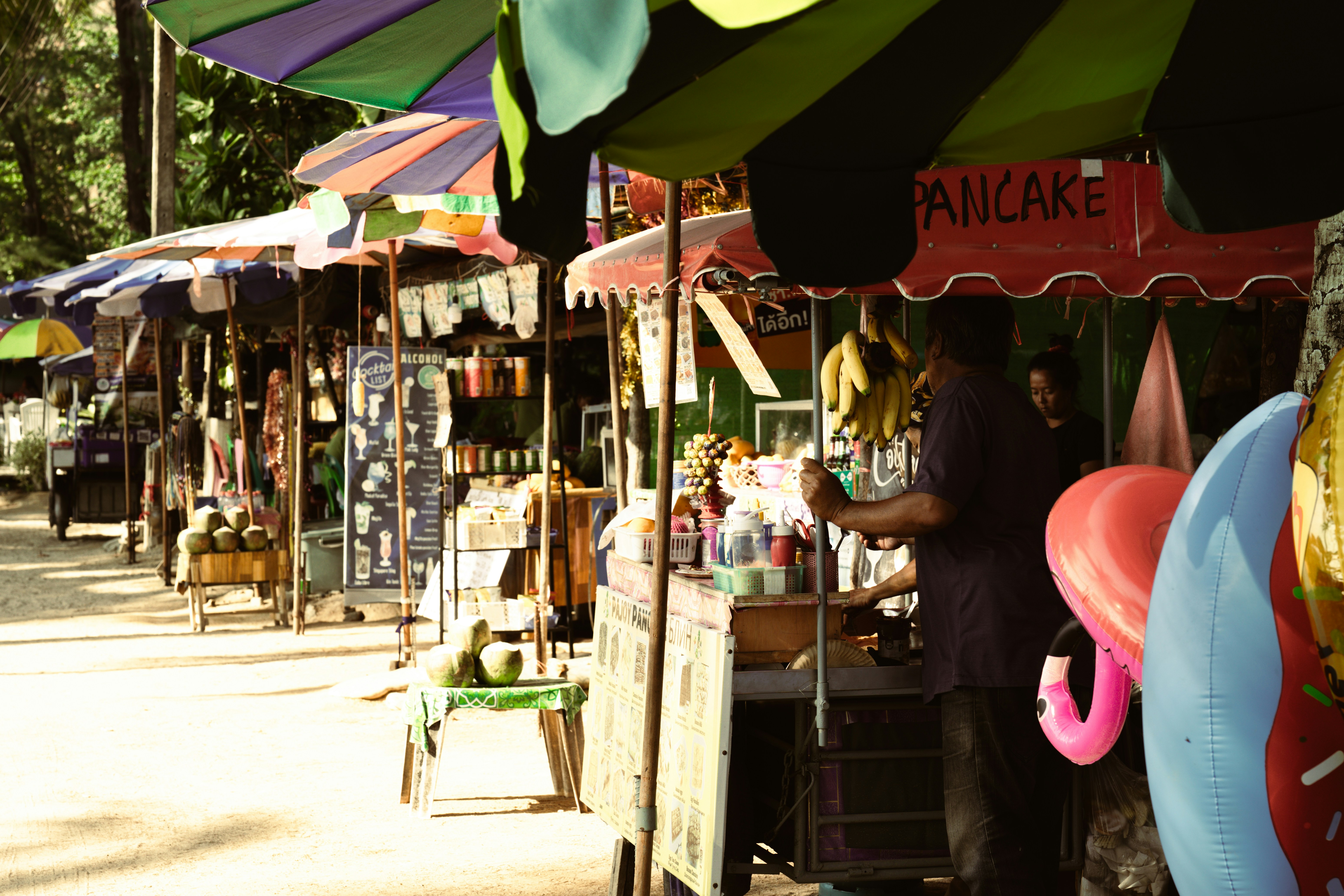 Street vendor stalls with colorful umbrellas and tropical fruits in a sunlit market.