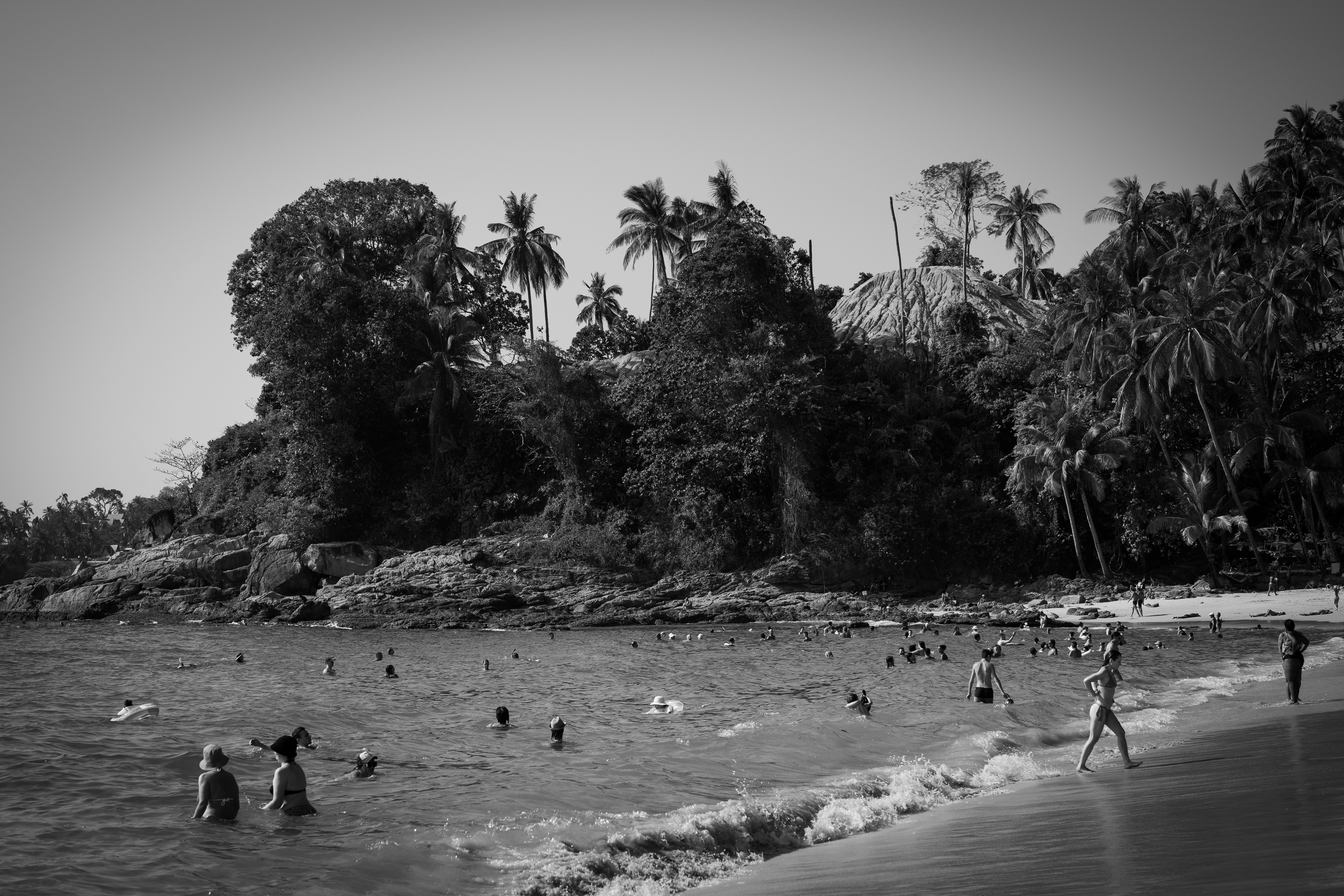 Beachgoers enjoy the ocean near a rocky, tree-covered cliff.