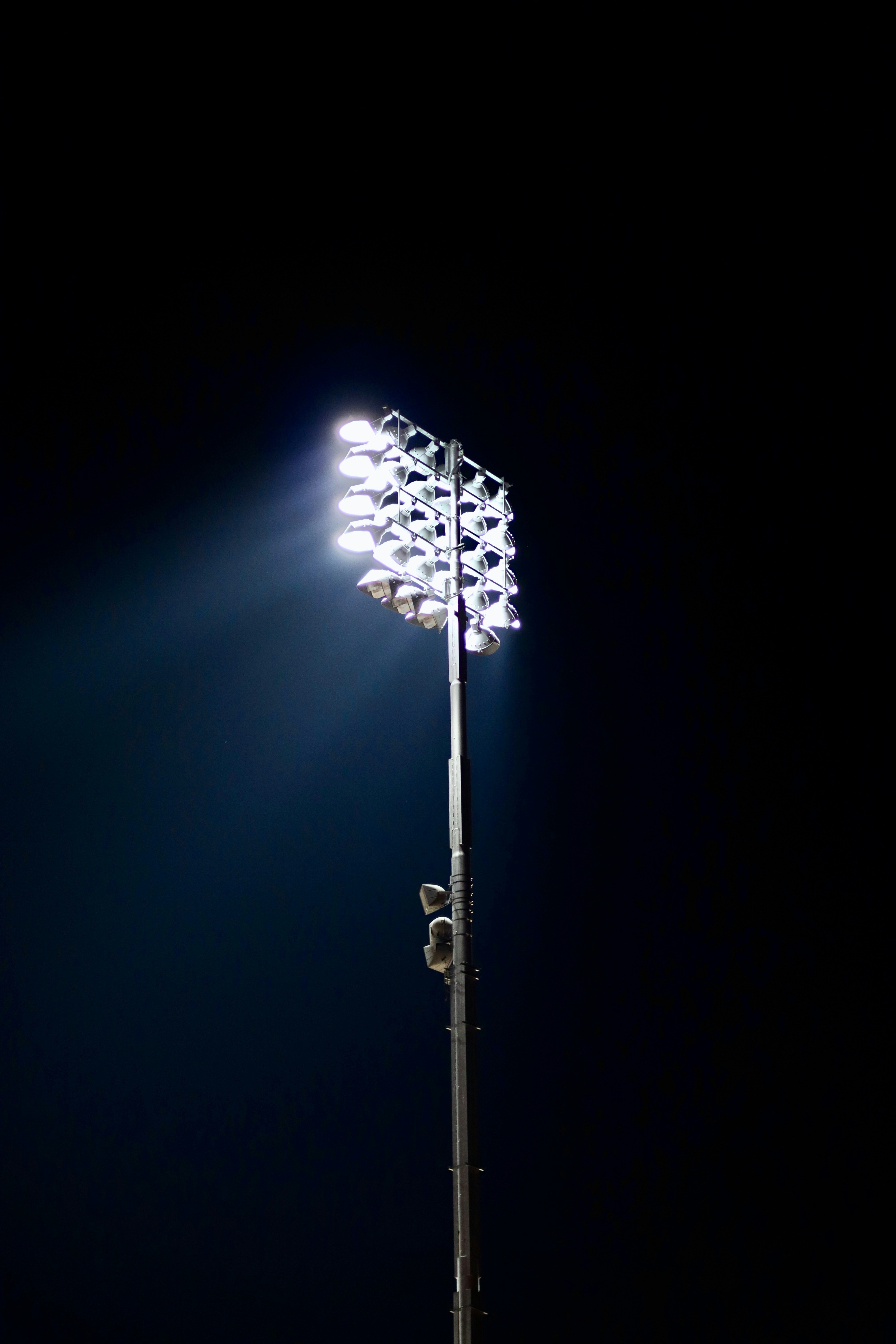 Night photograph of a tall floodlight tower with a bright, multi-lamp array piercing the dark sky. The frame emphasizes the solitary structure and its glow against expansive negative space.