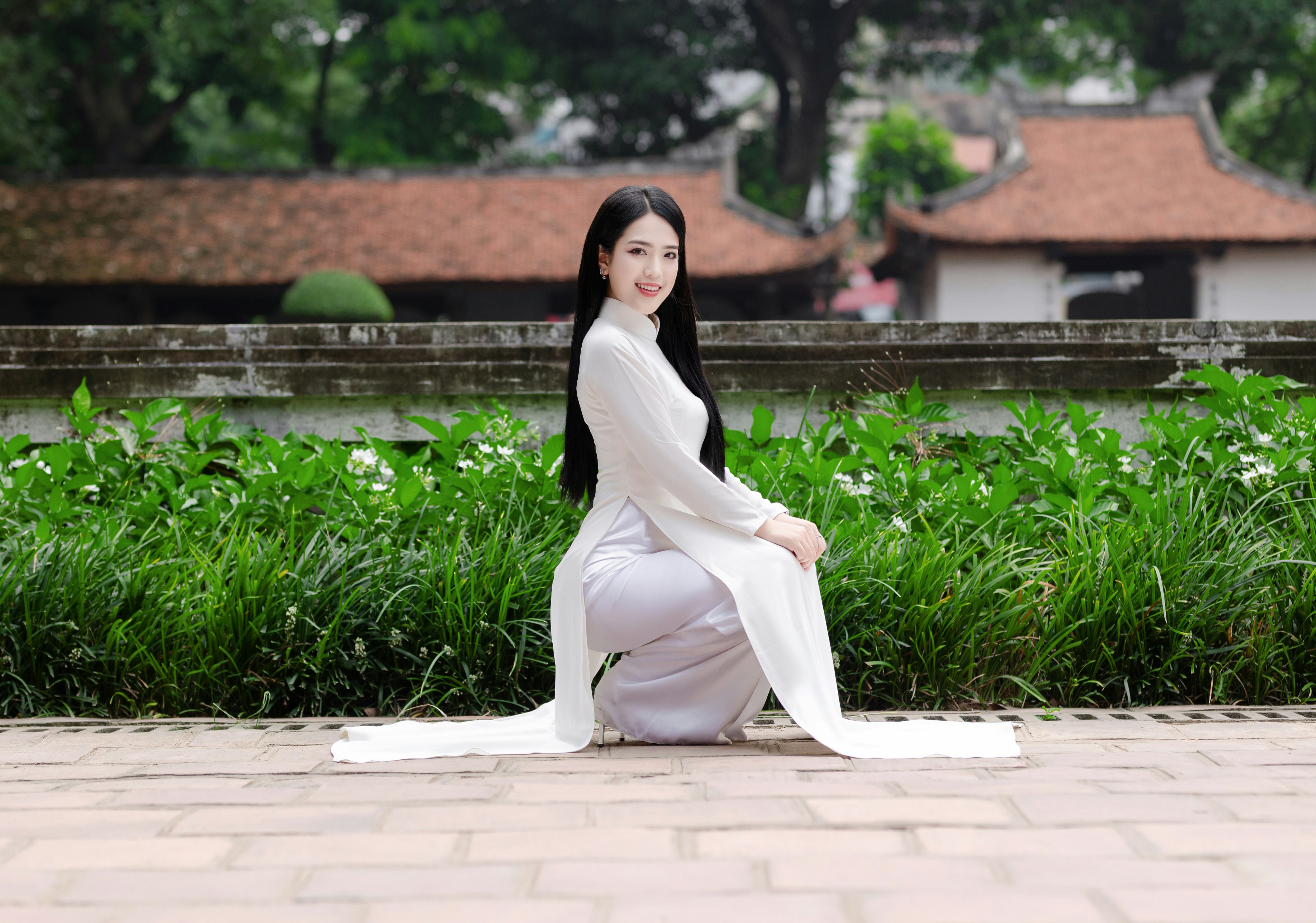 Woman in white Ao Dai kneeling by lush greenery with traditional architecture in the background.