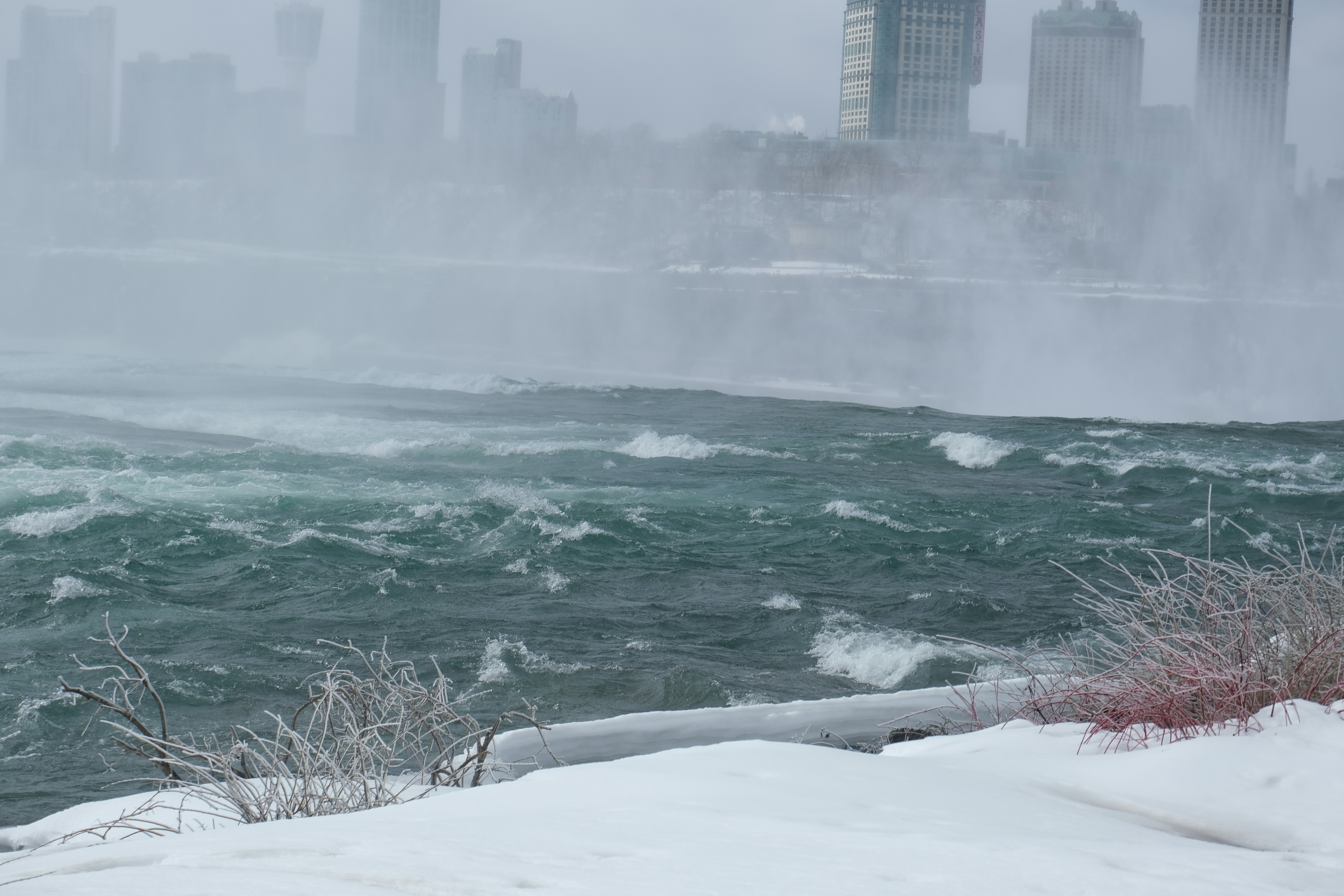 Choppy waters meet a snowy shoreline under a misty city skyline.