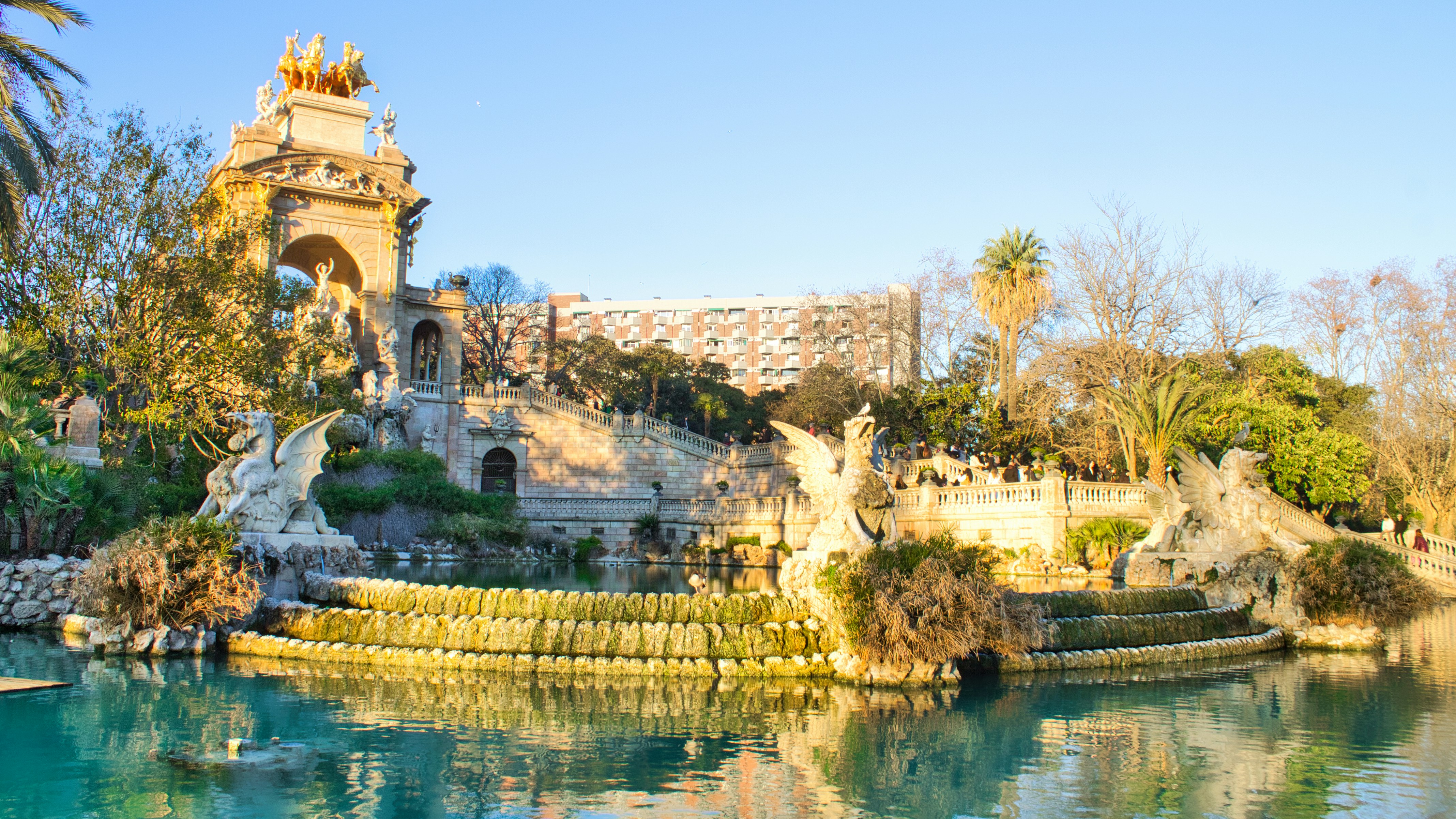Elaborate stone fountain with mythical sculptures surrounded by lush greenery and a reflective pond.