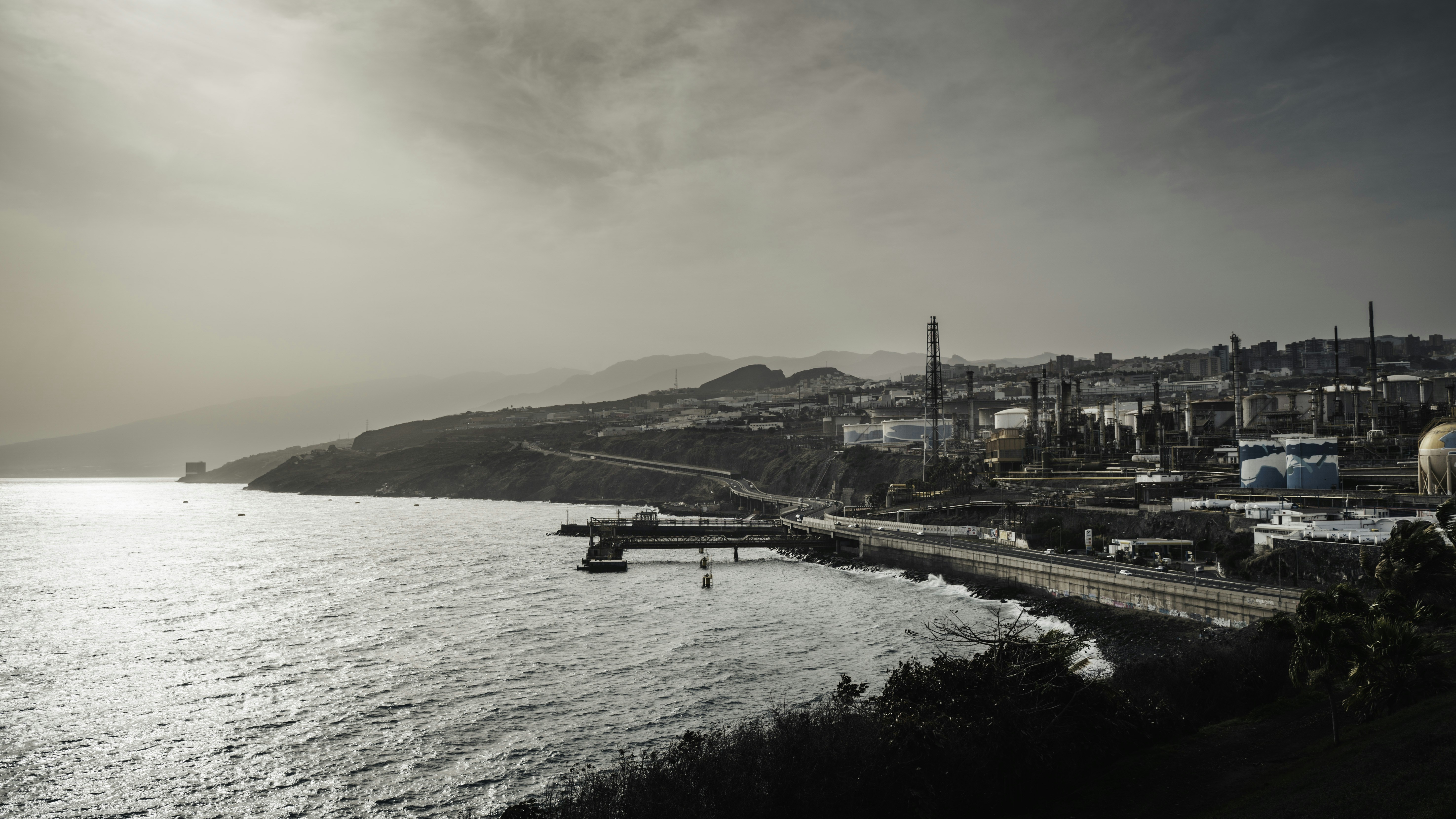 Industrial coastline with factories and smokestacks under a cloudy sky, with the ocean reflecting muted sunlight.