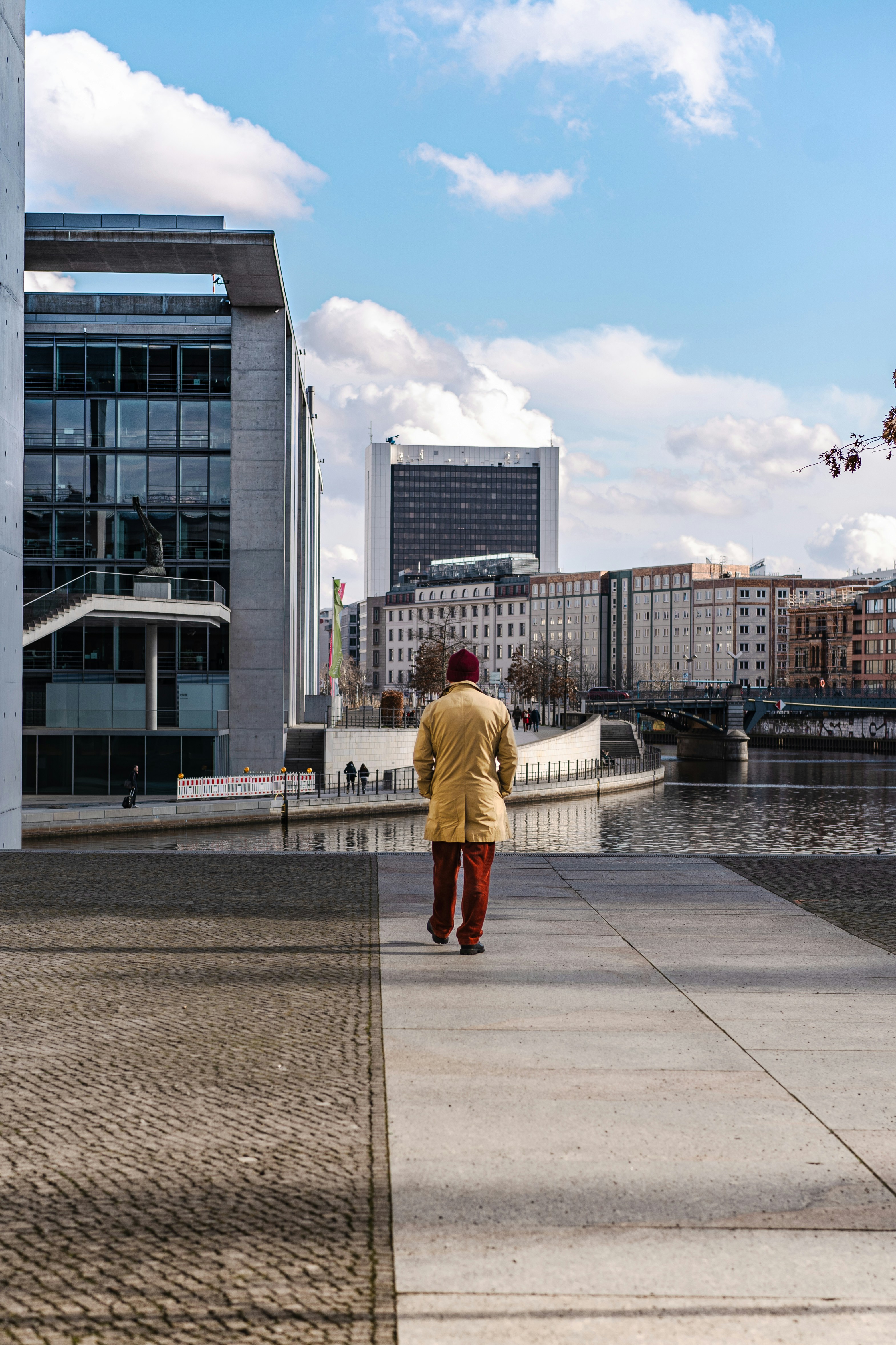 Mustard Coat, Red Trousers: A Solitary Stroll Along a Glass-Fronted Riverside Cityscape