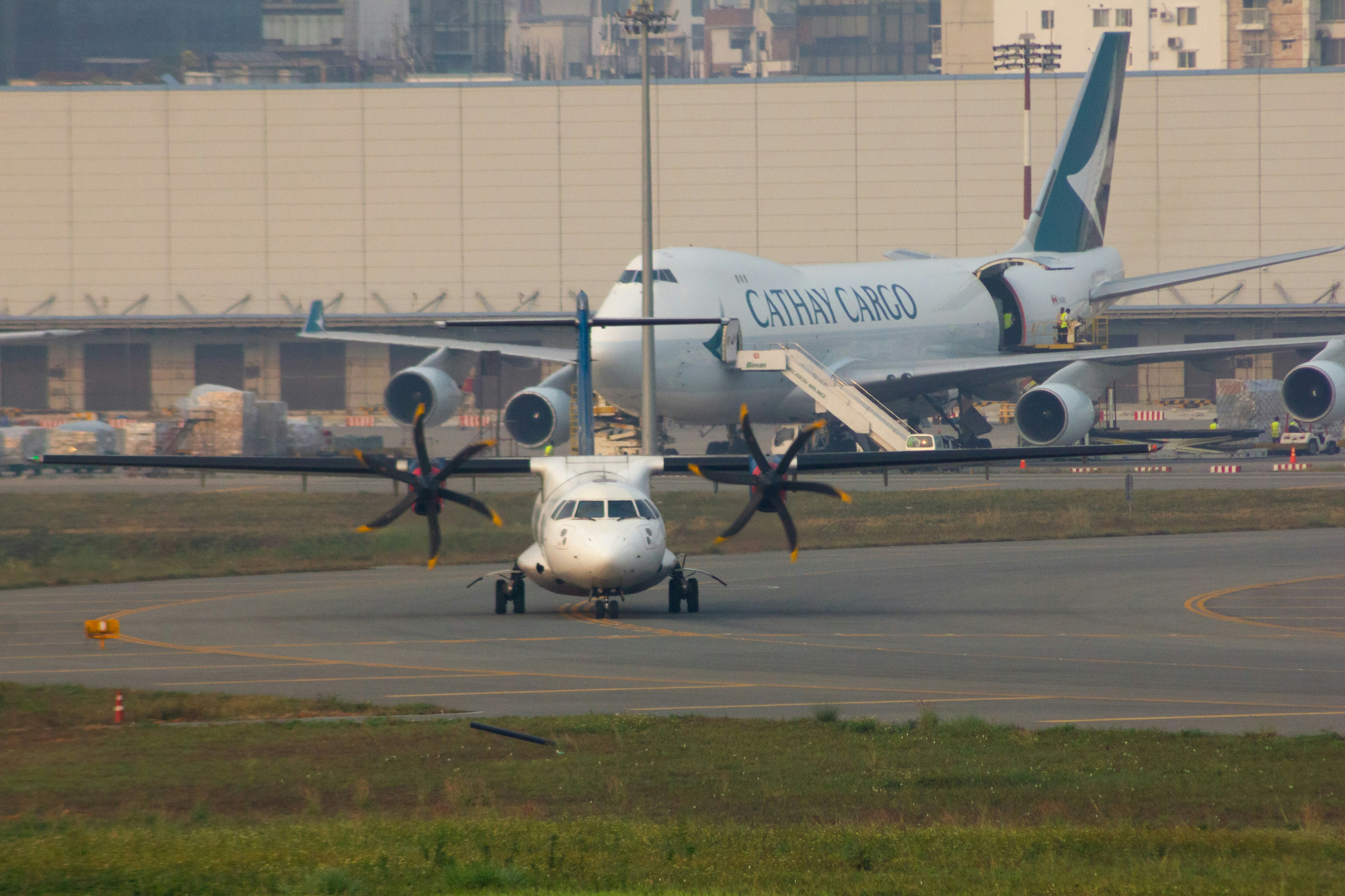 A Cathay cargo plane unloads while a smaller turboprop aircraft waits on the runway.