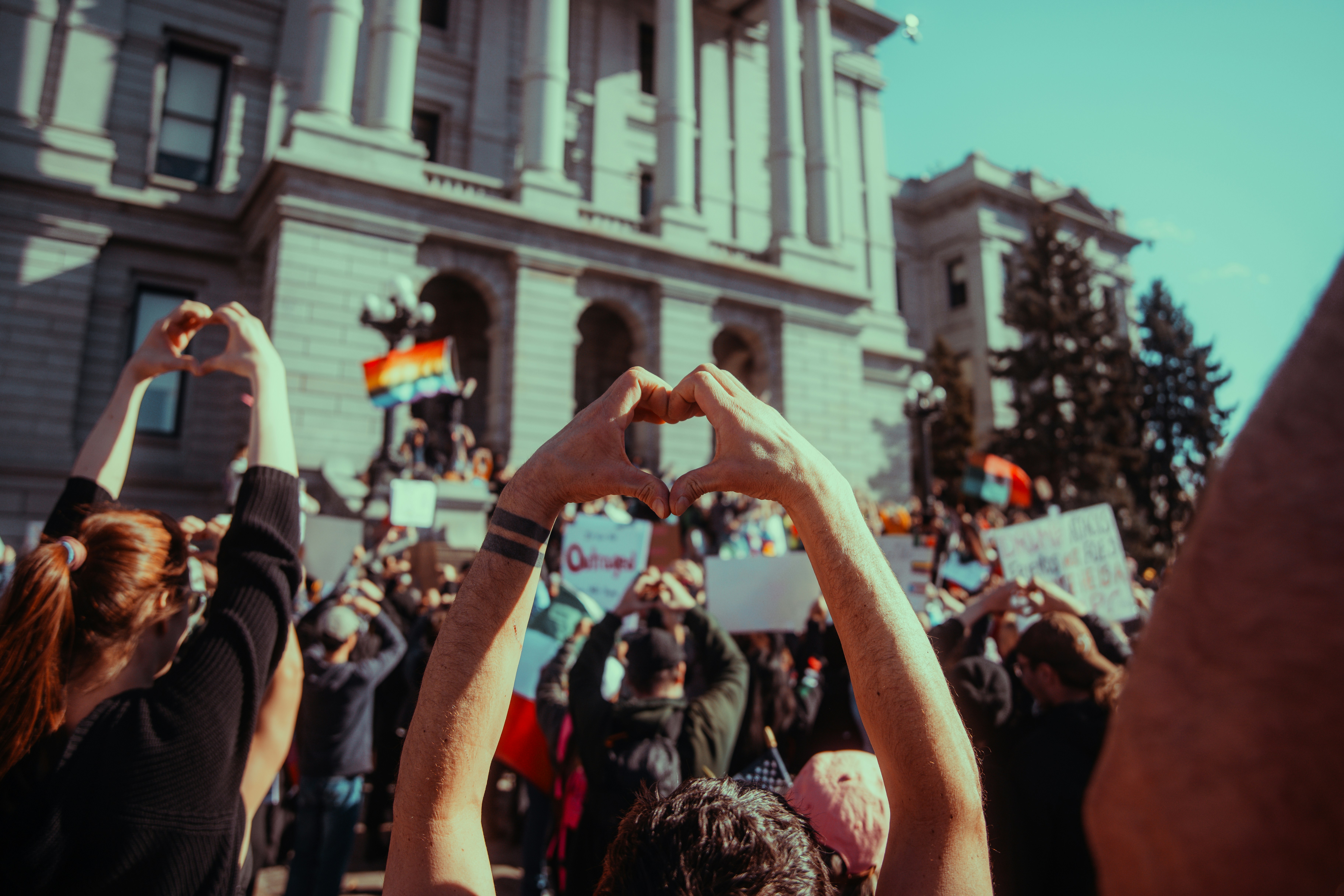 Crowd outside a historic building with raised hands forming heart shapes amidst protest signs and flags.