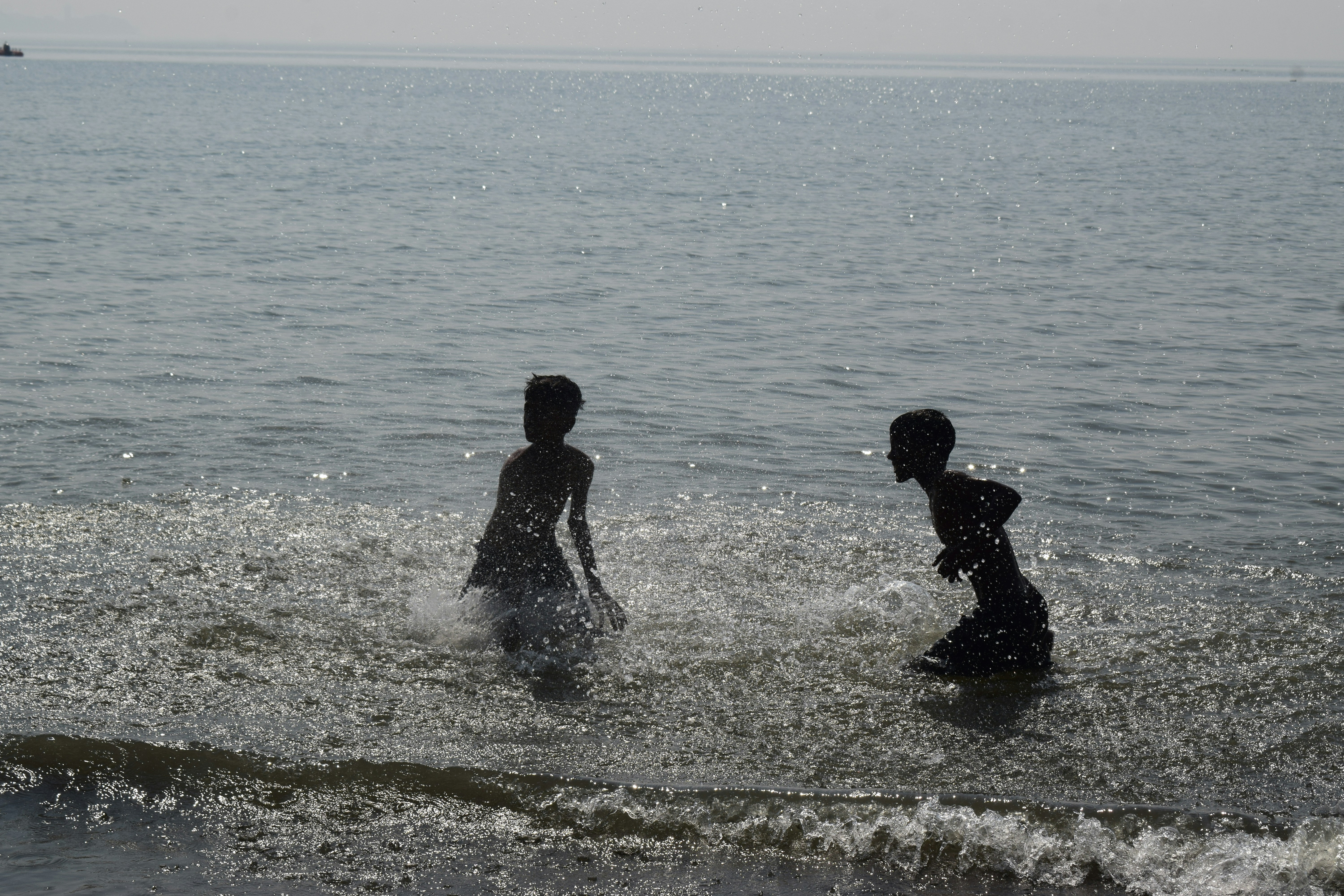 Two children play in the shimmering waves of the sea, silhouetted against the bright sky.