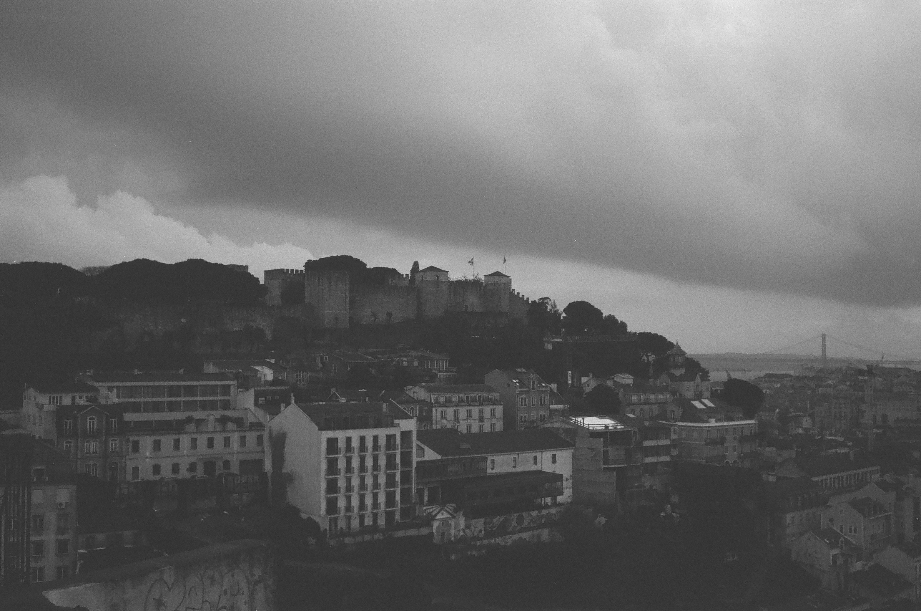 Overcast sky looms above a historic fortress and cityscape, with a distant bridge visible through the mist.