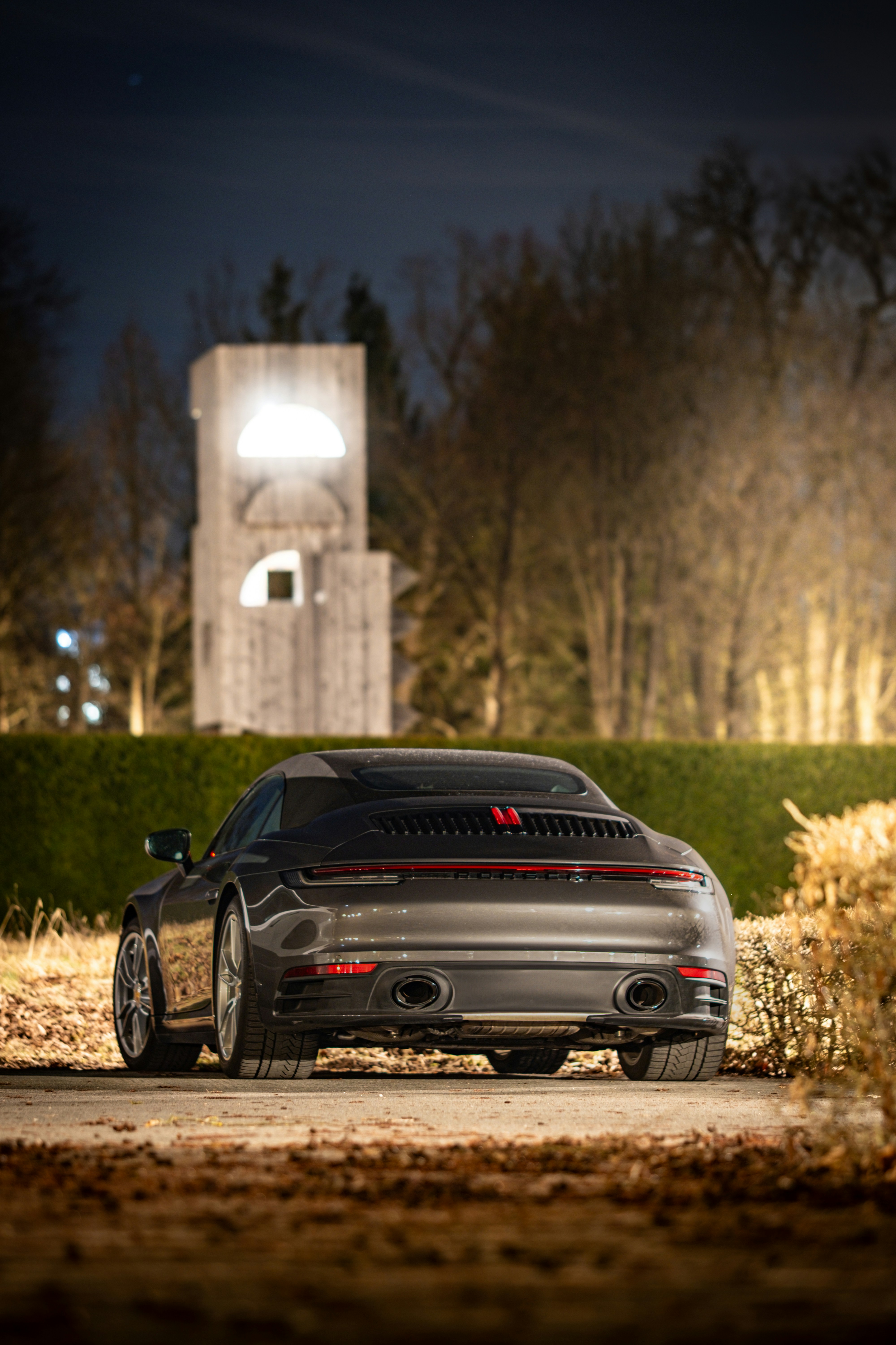 Rear view of a Porsche 992 parked on a leaf-strewn path with a softly lit tower and trees in the background.