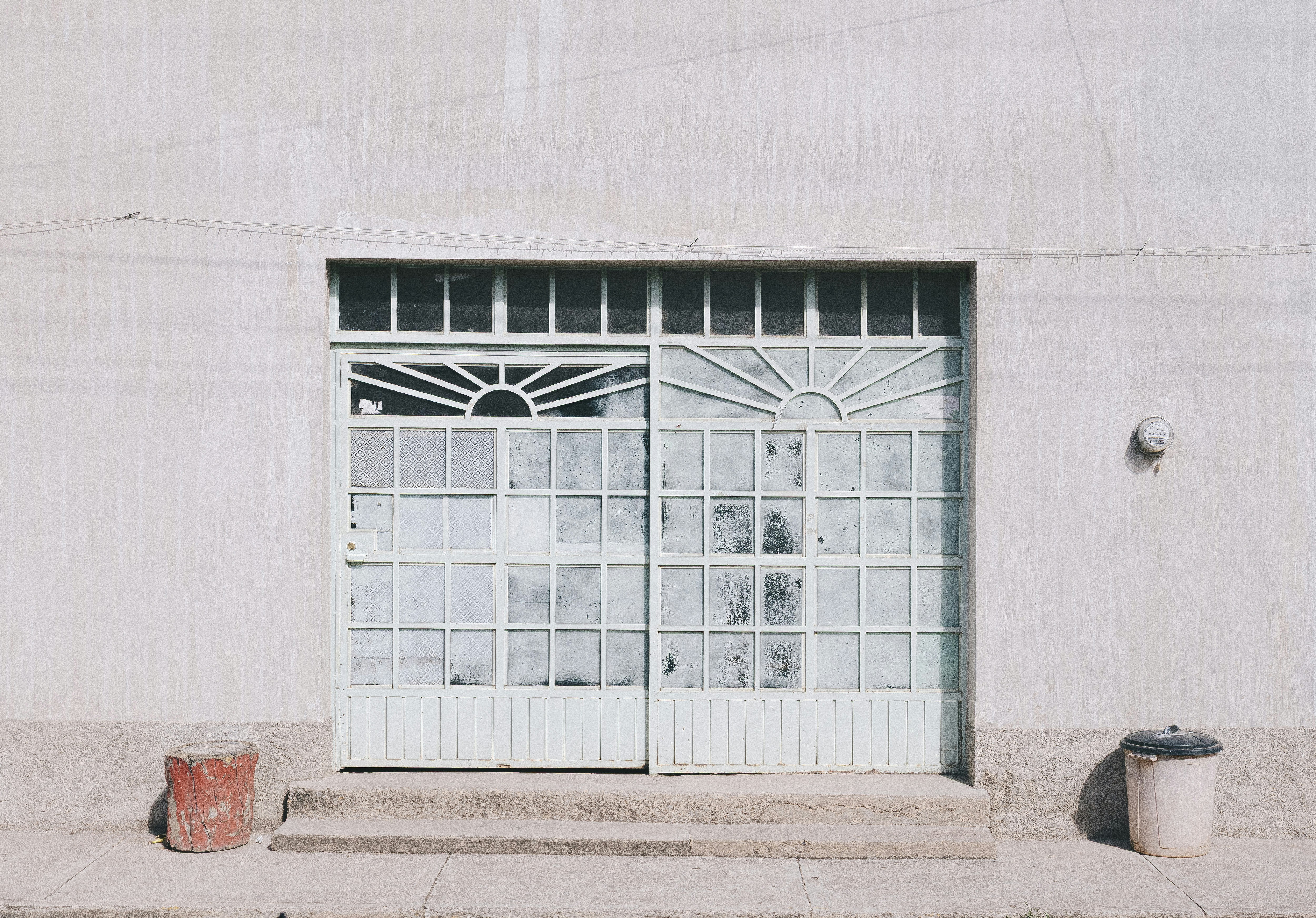 Pale blue double doors with geometric patterns, flanked by a red stool and a trash bin on a sunlit sidewalk.