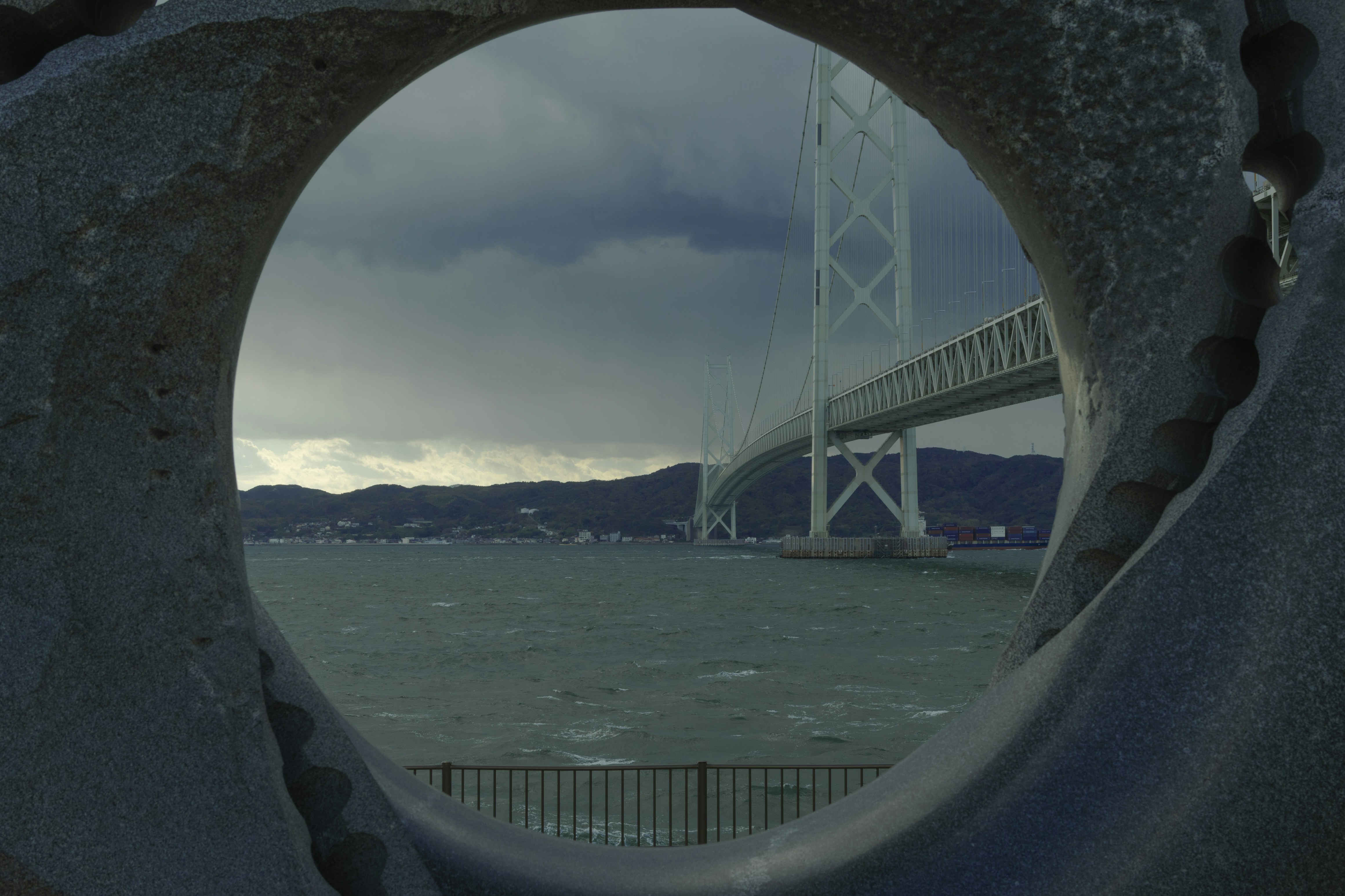 Circular stone sculpture frames a distant suspension bridge over a turbulent sea under moody skies.