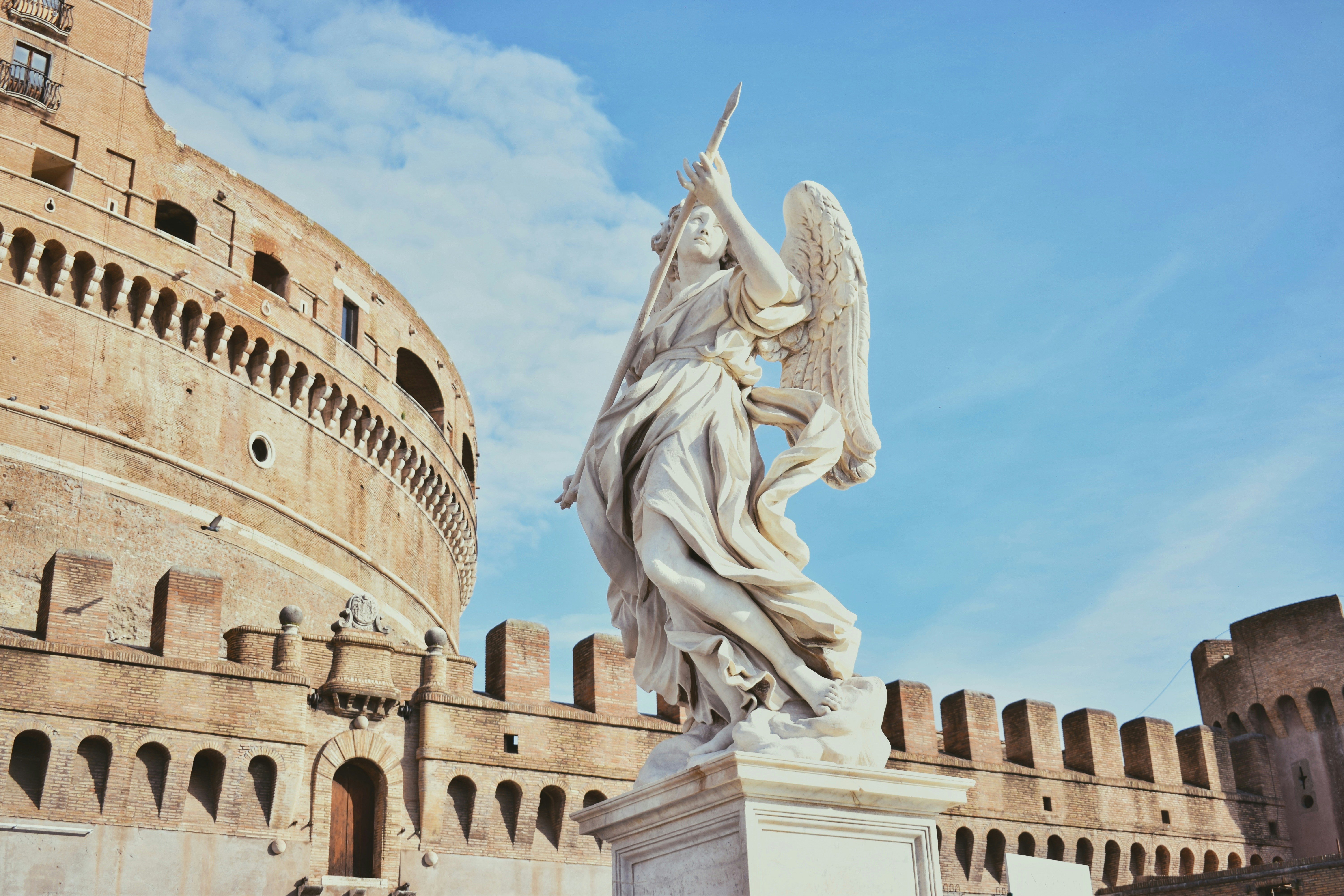Marble angel statue holding a spear stands in front of the historic Castel Sant'Angelo under a clear sky.