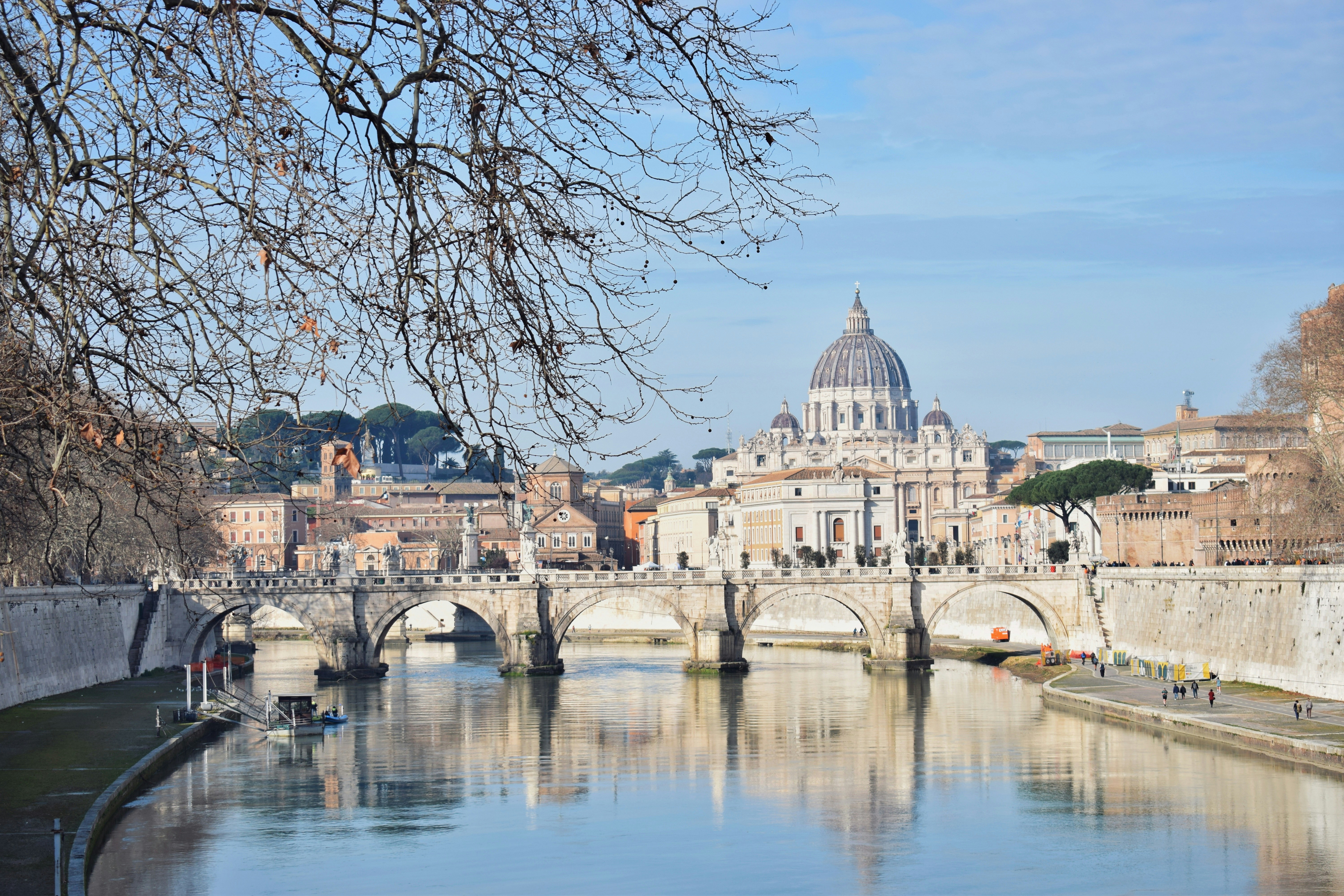 St. Peter's Basilica and Sant'Angelo Bridge reflected in the calm waters of the Tiber River under a clear blue sky.