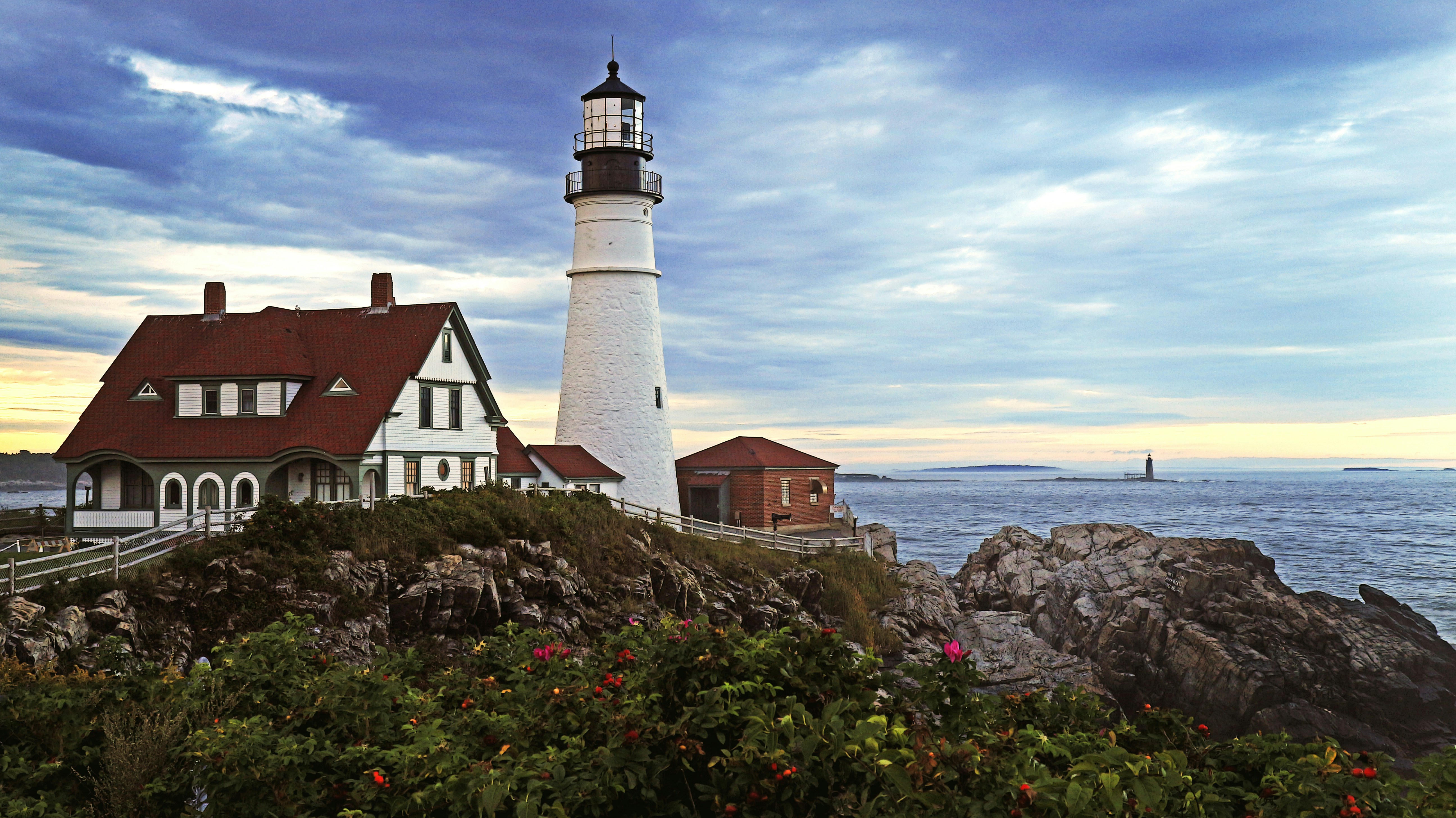 White lighthouse and red-roofed house perched on rocky coastline with a vibrant sky and distant ocean horizon.
