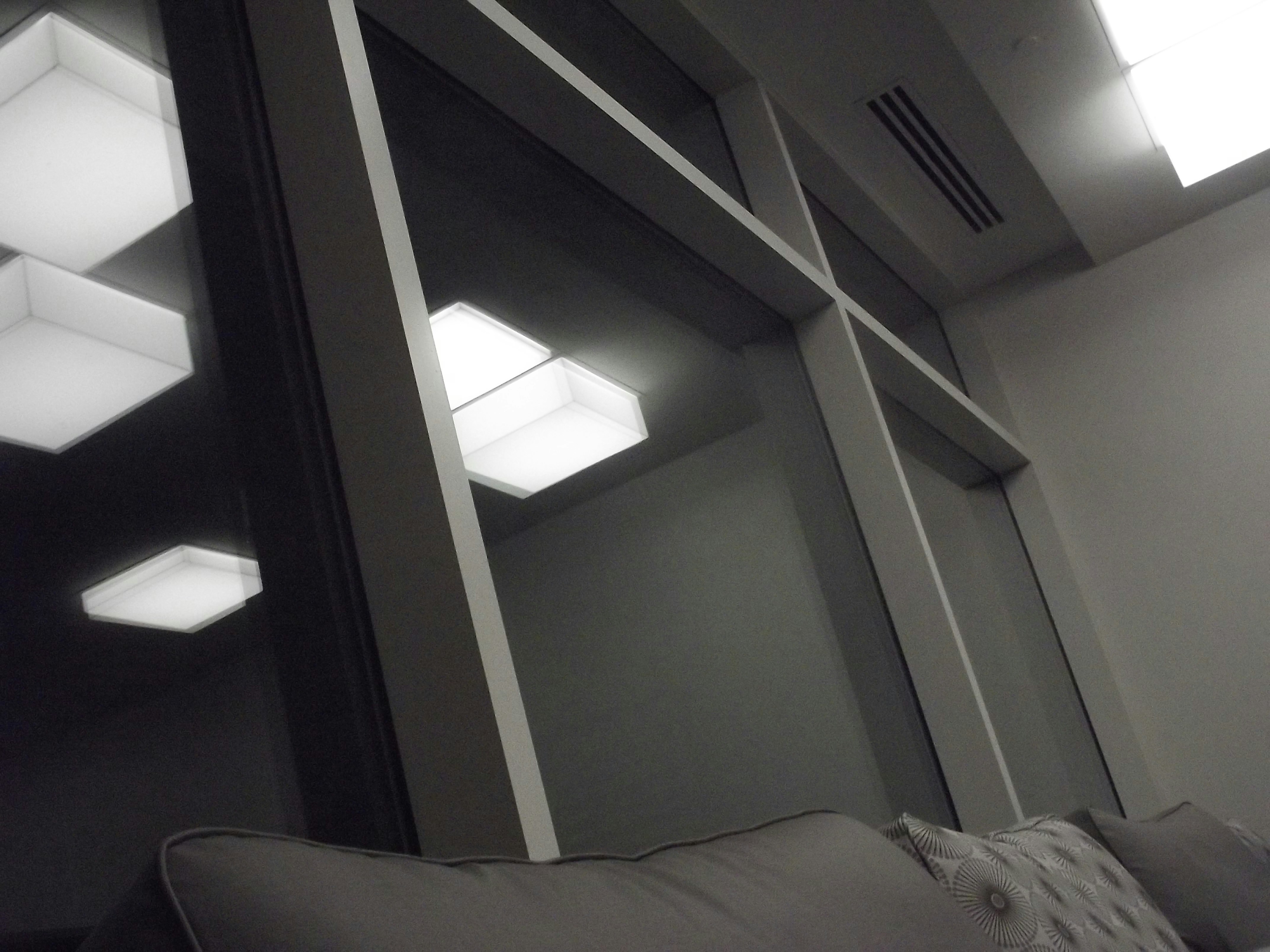 Geometric light fixtures reflect off glass panels in a contemporary interior space, showcasing a blend of light and shadow.