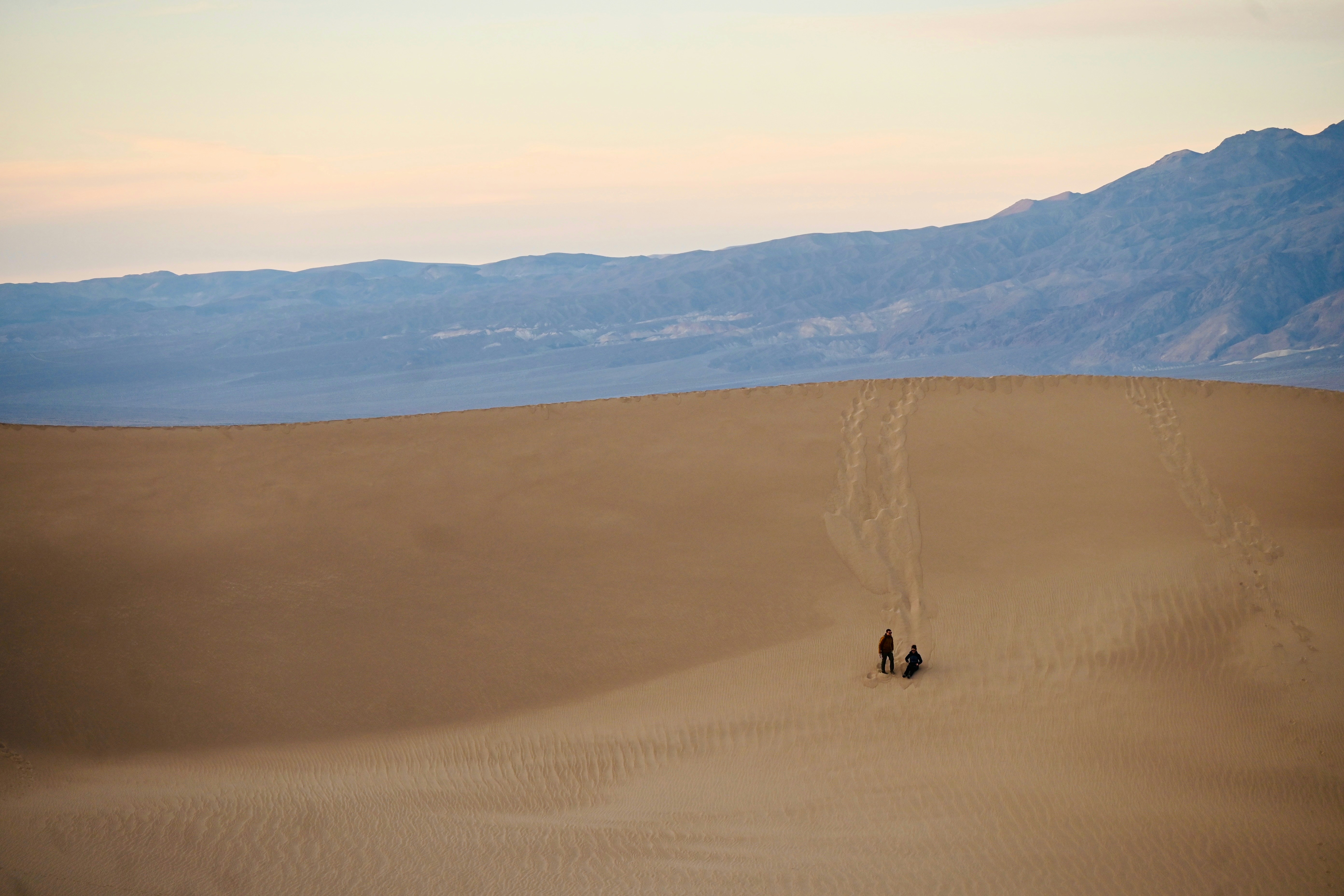 Two figures rest on a vast sand dune under a pastel sky with distant mountains.
