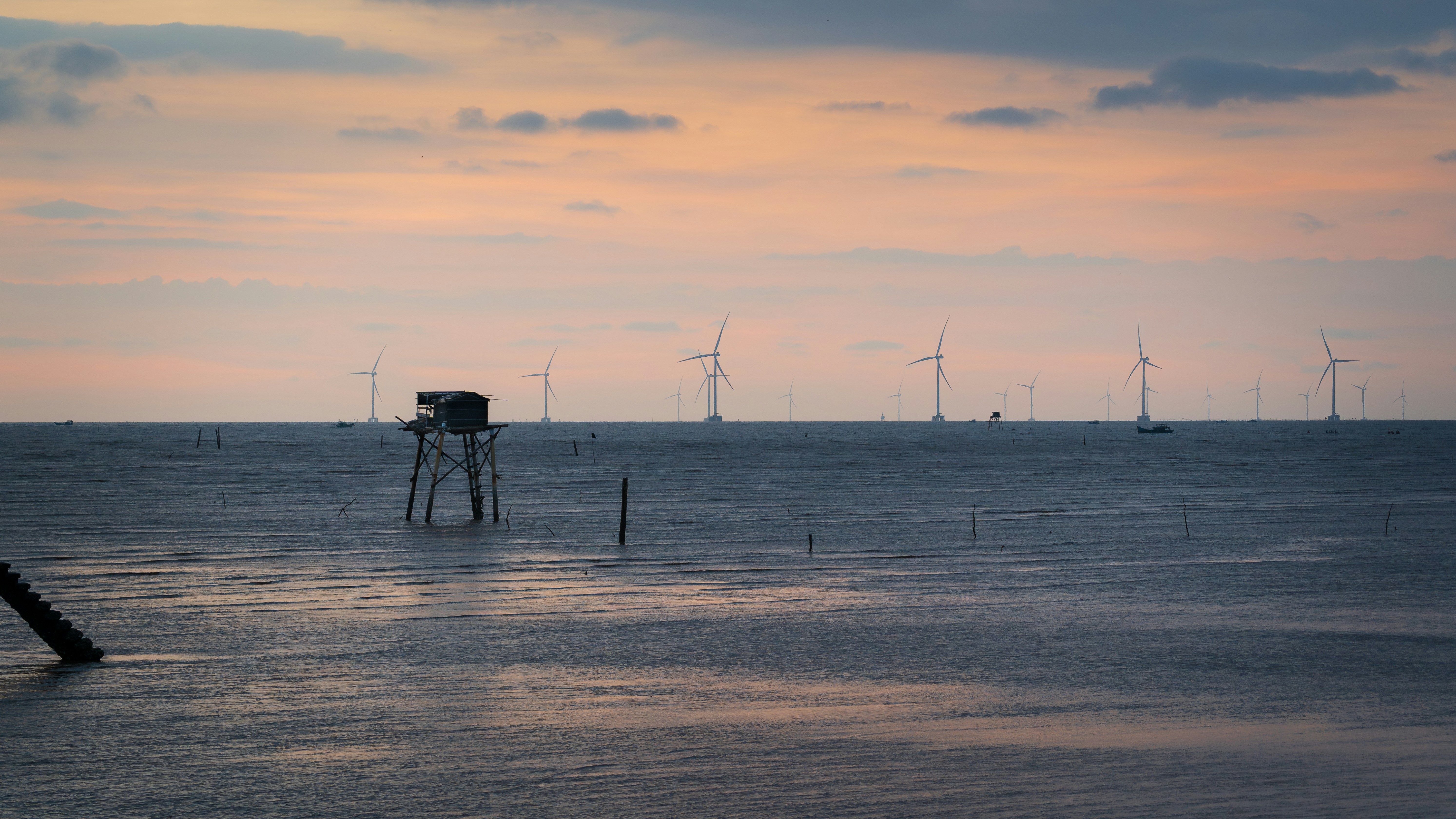 Wind turbines stand tall against a pastel sunset over calm waters near an oyster farm in Vietnam.