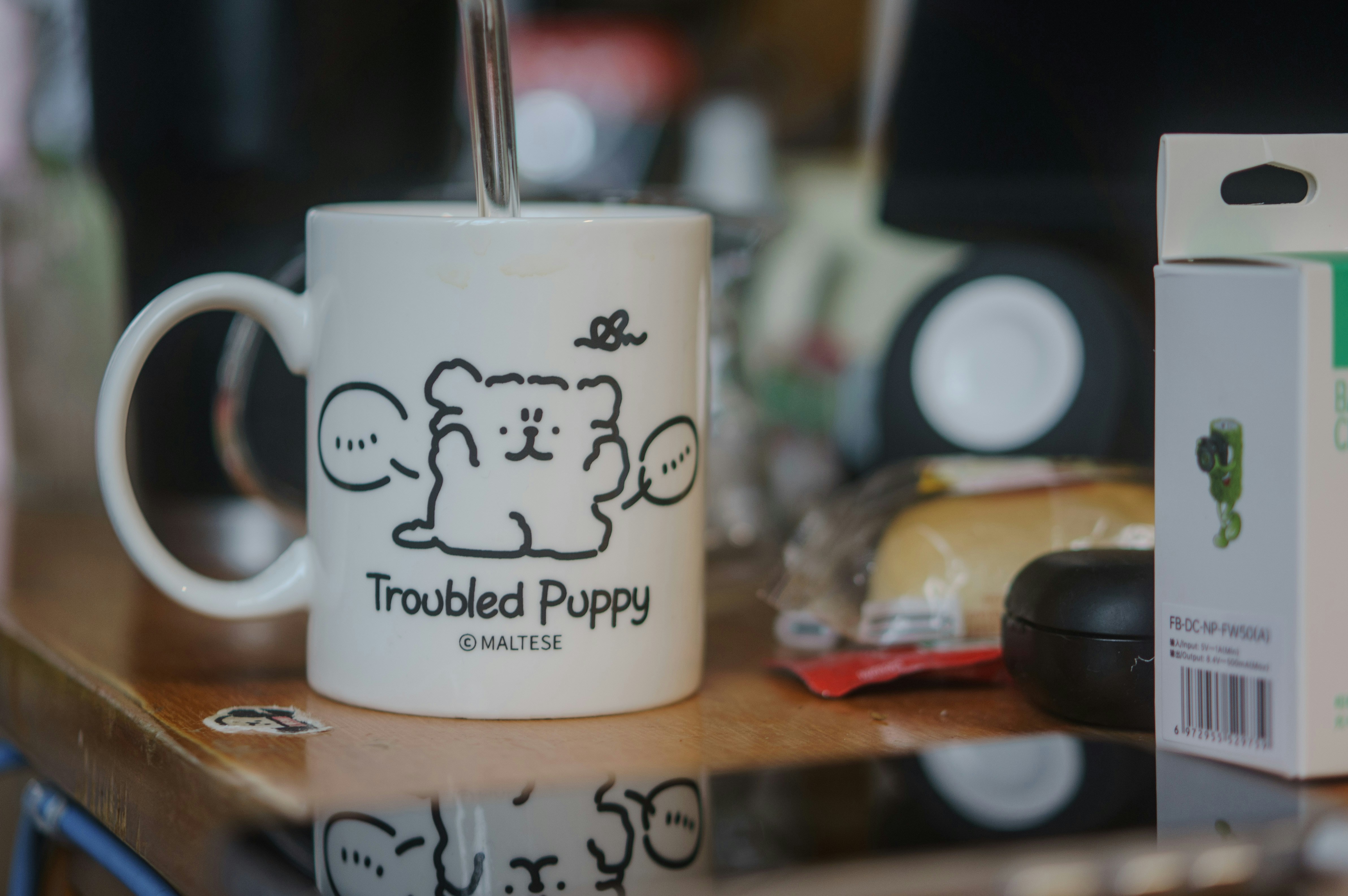 Close-up of a white mug bearing a 'Troubled Puppy' Maltese illustration on a glossy desk, with blurred desk items in the background.