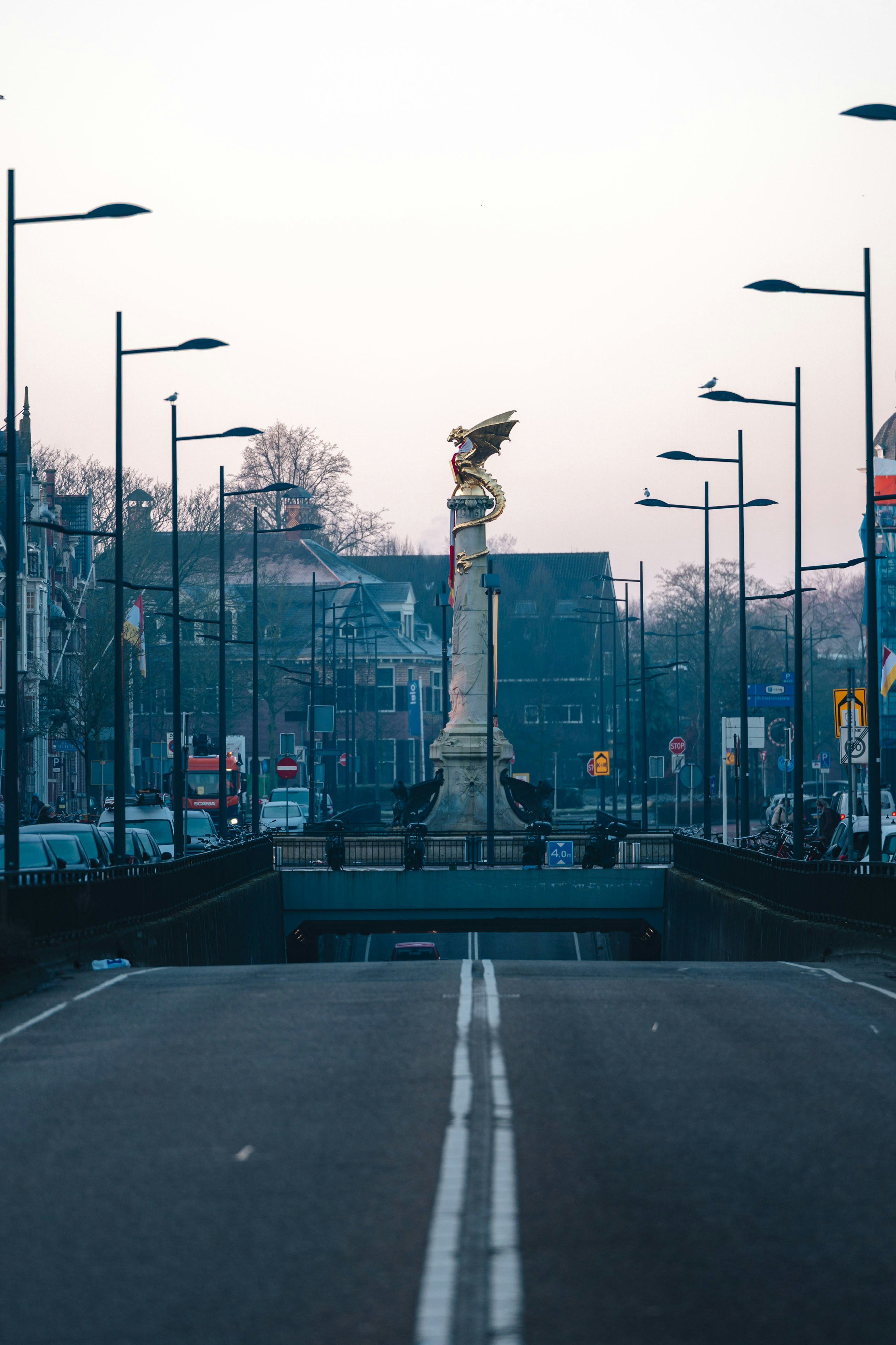 Statue with wings in the center of a quiet city street at dawn, framed by streetlights and buildings.