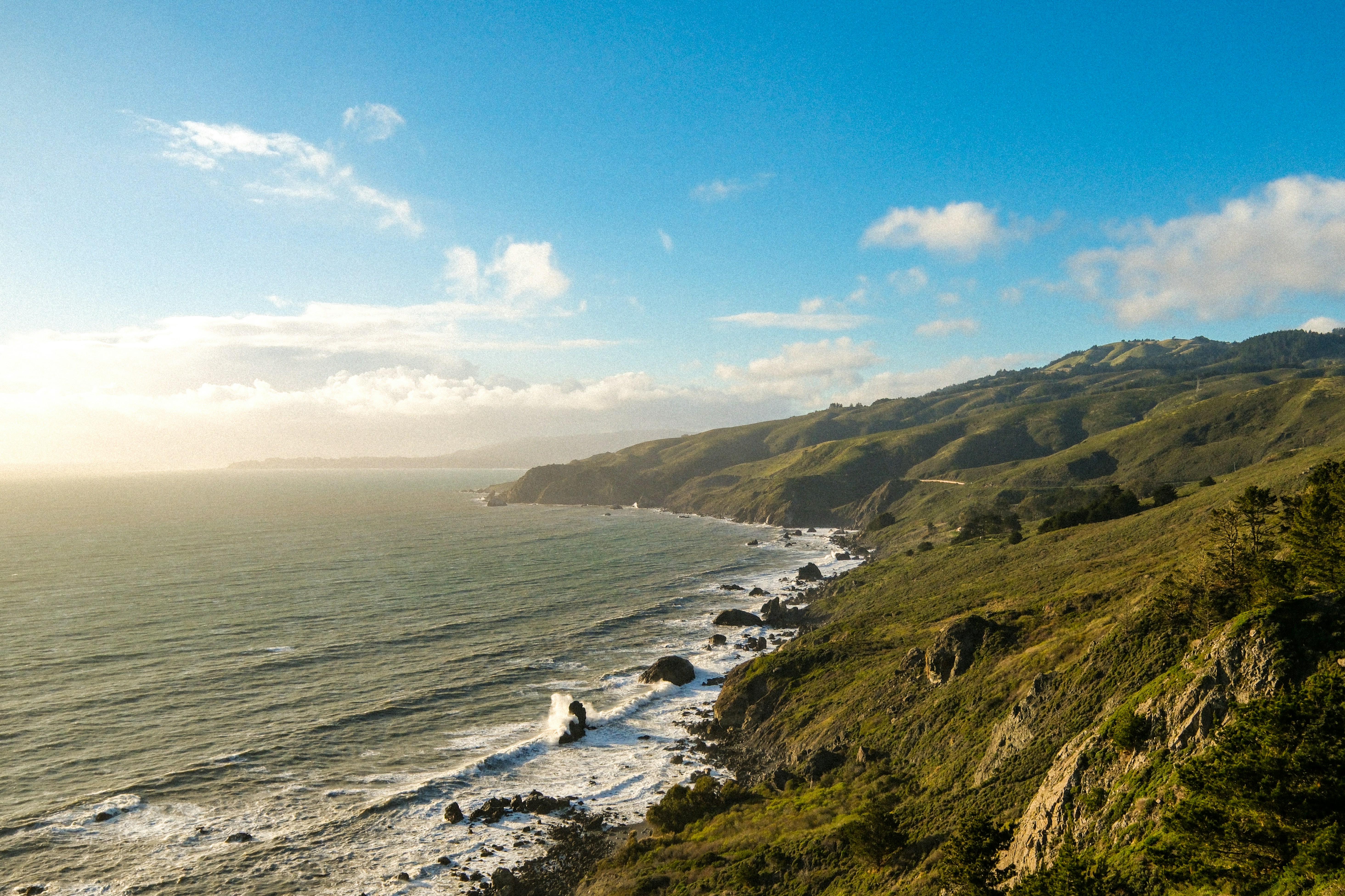 Rocky coastline with waves crashing under a clear blue sky and rolling green hills.