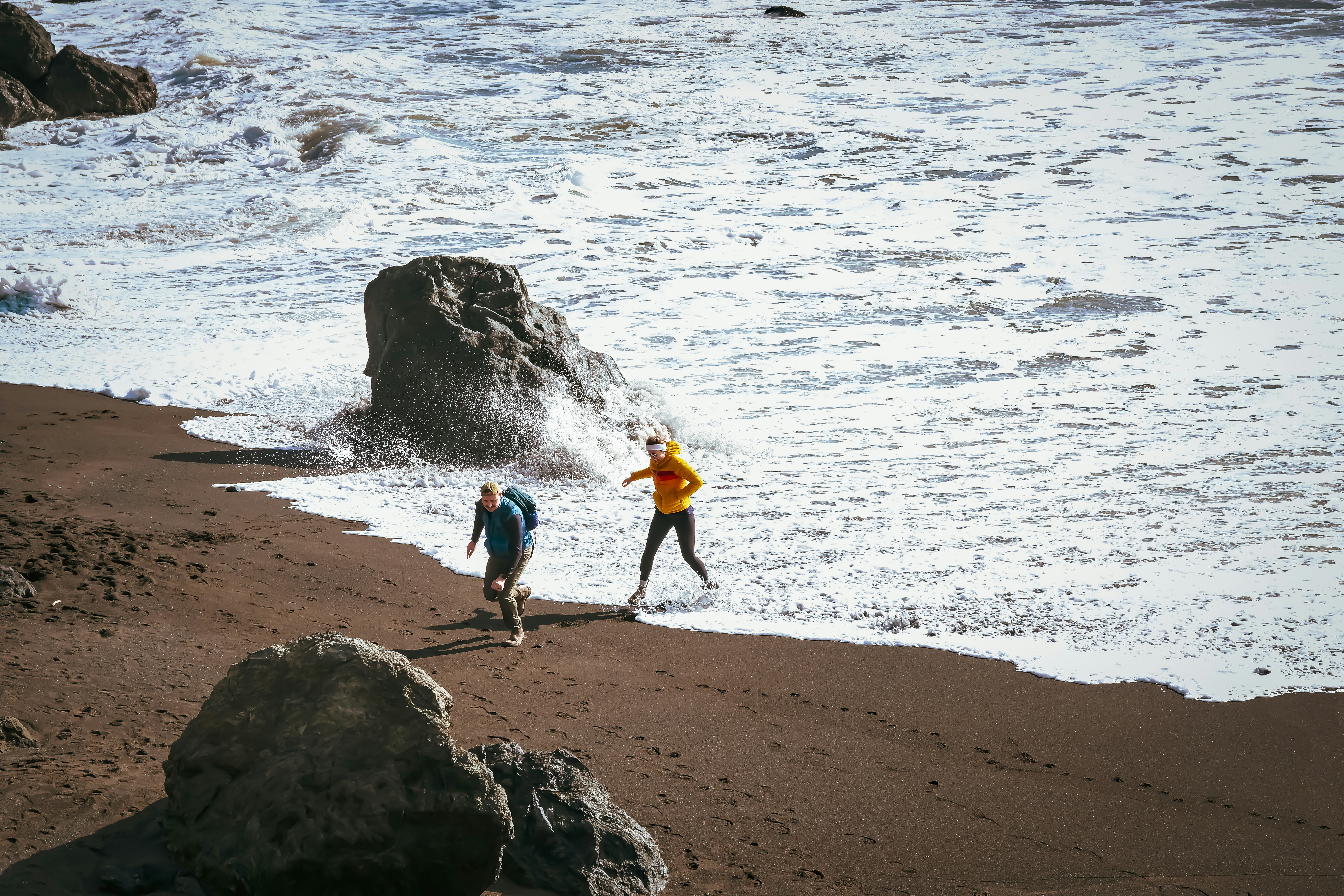Two people run along a sandy beach as waves crash against large rocks.