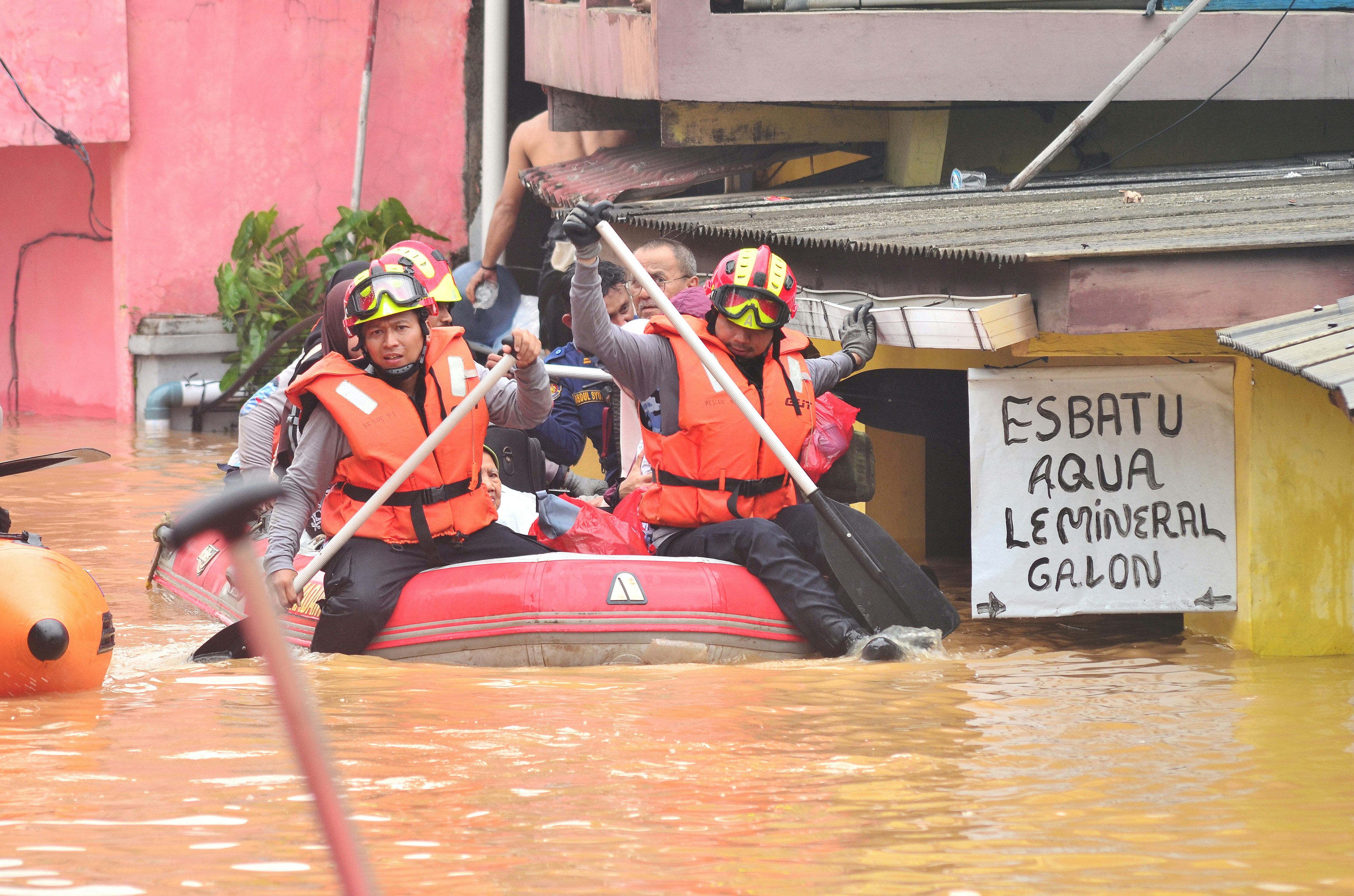 Rescue team evacuating people with an inflatable boat through a flooded urban area, wearing bright orange life vests.