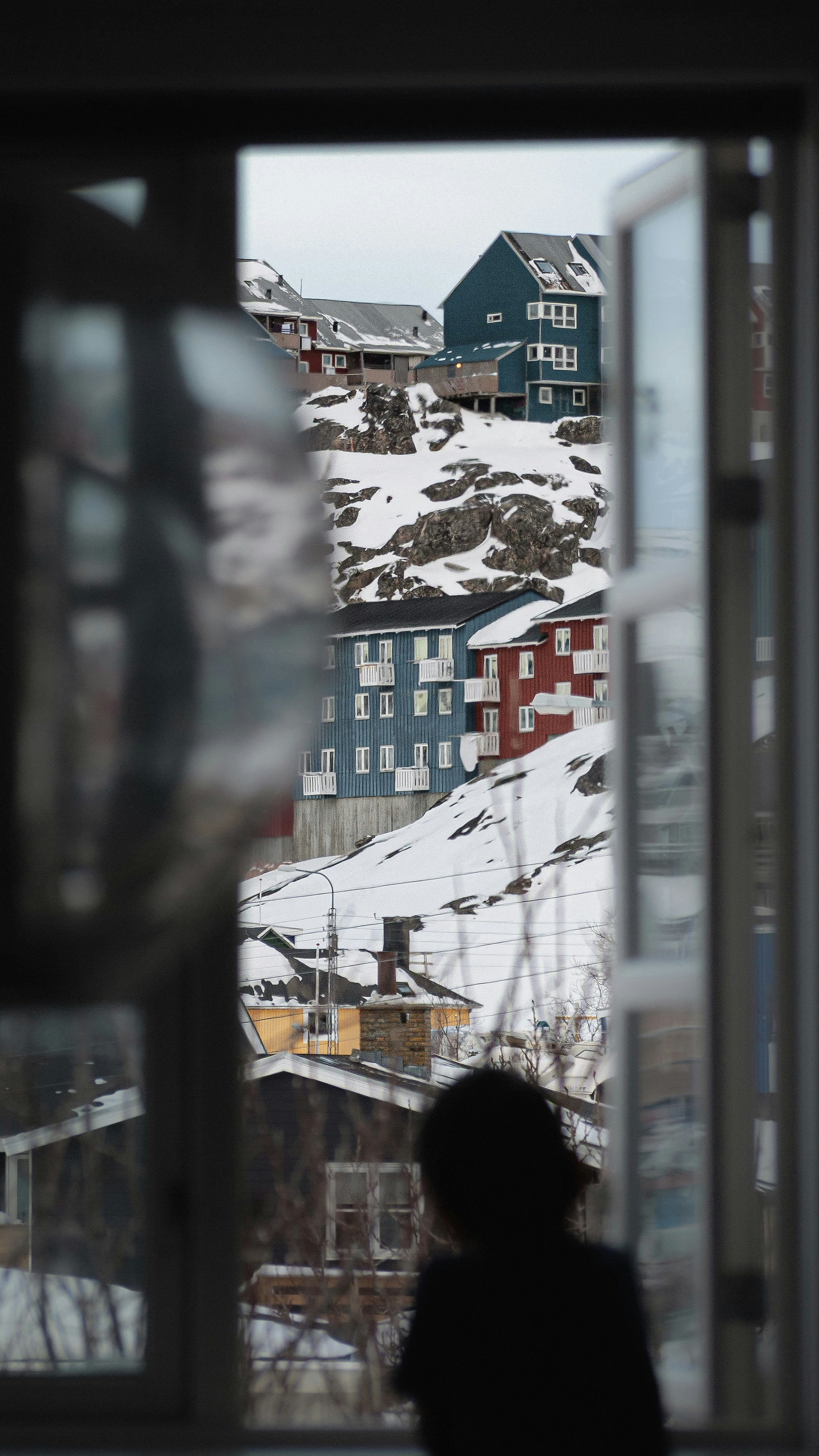 Colorful hillside houses in snow viewed through a narrow window, with a dark silhouette in the foreground.