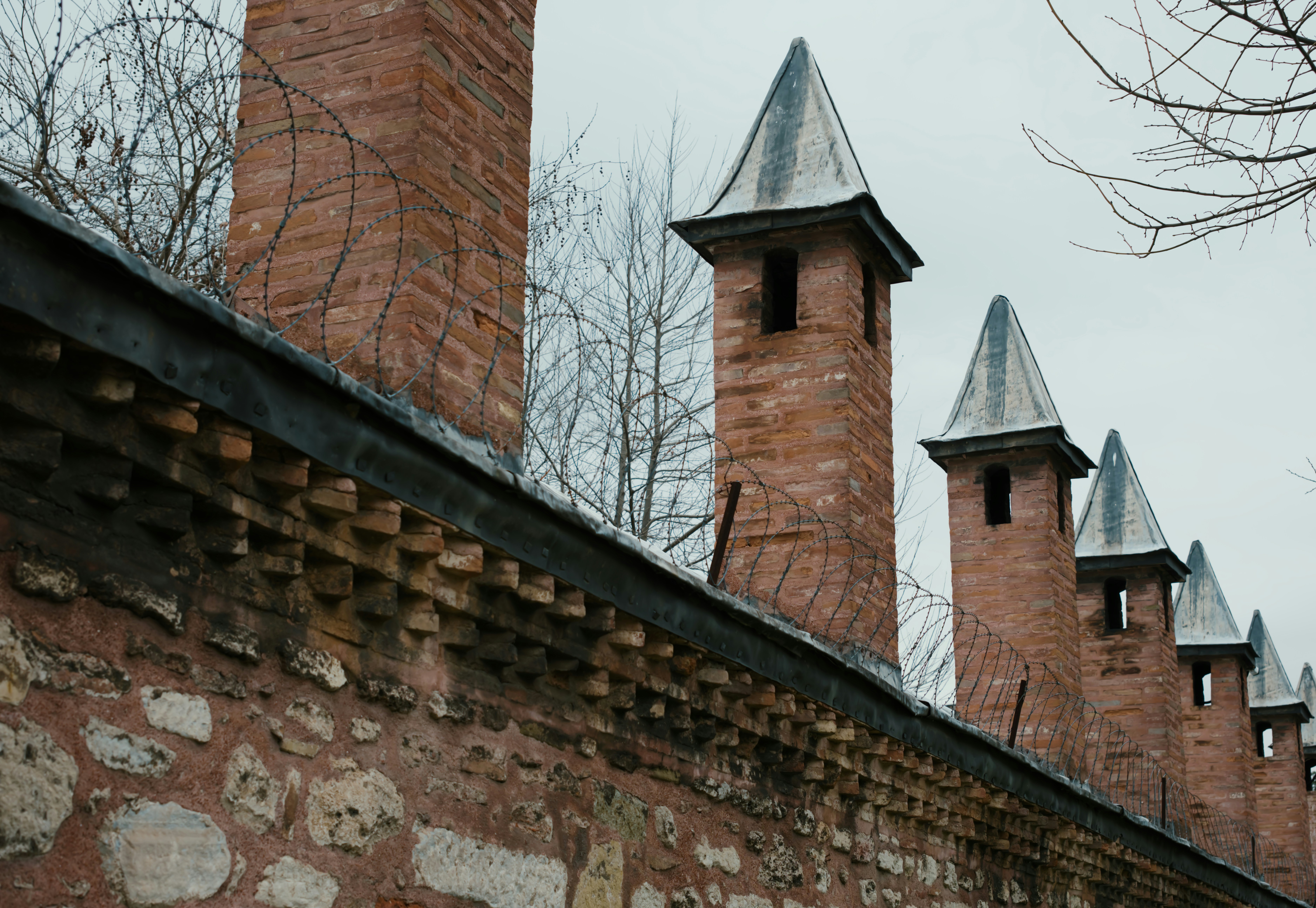 Brick towers with metal roofs and barbed wire atop a stone wall under a cloudy sky.