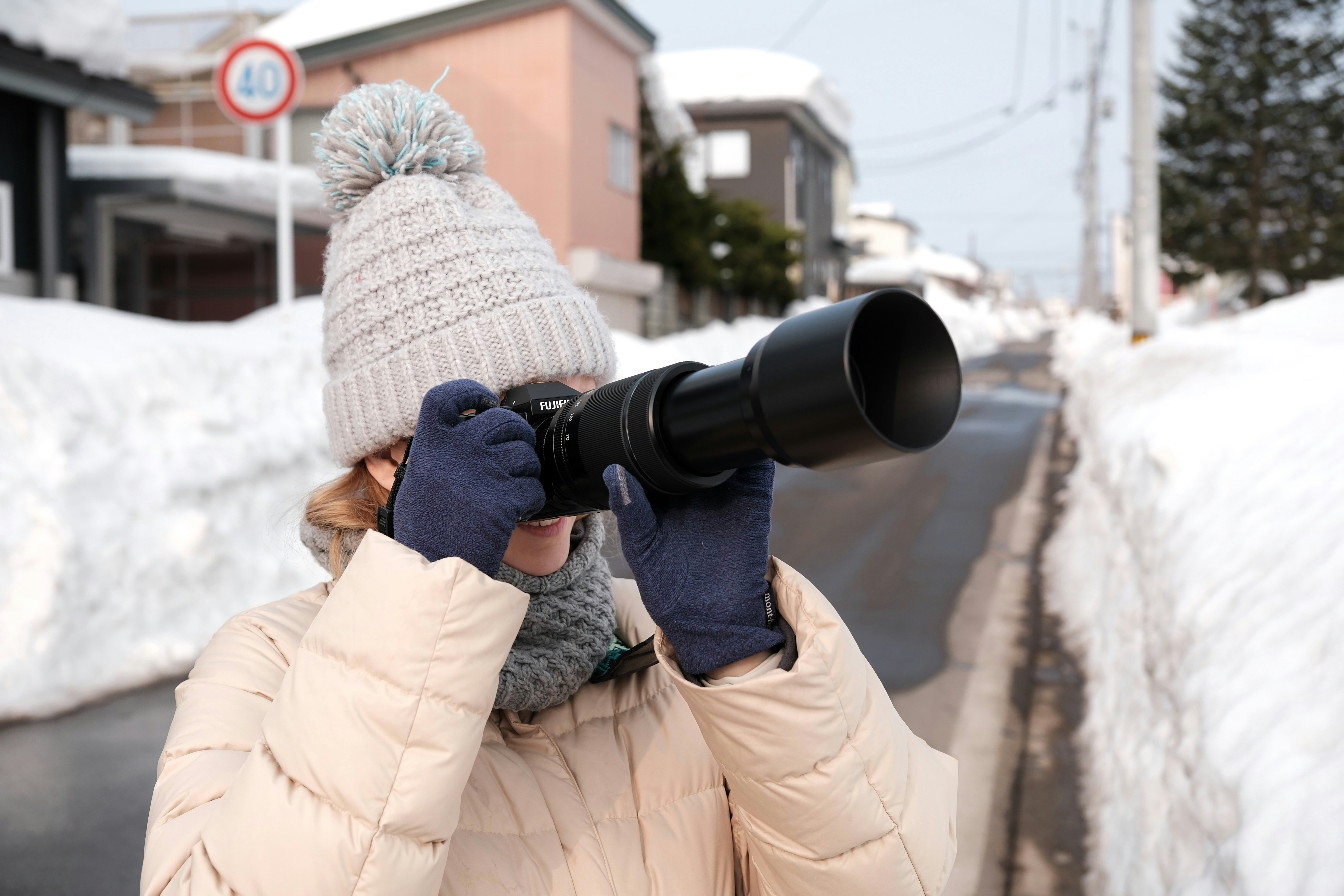 Person in winter attire photographing a snowy urban street with a long lens.