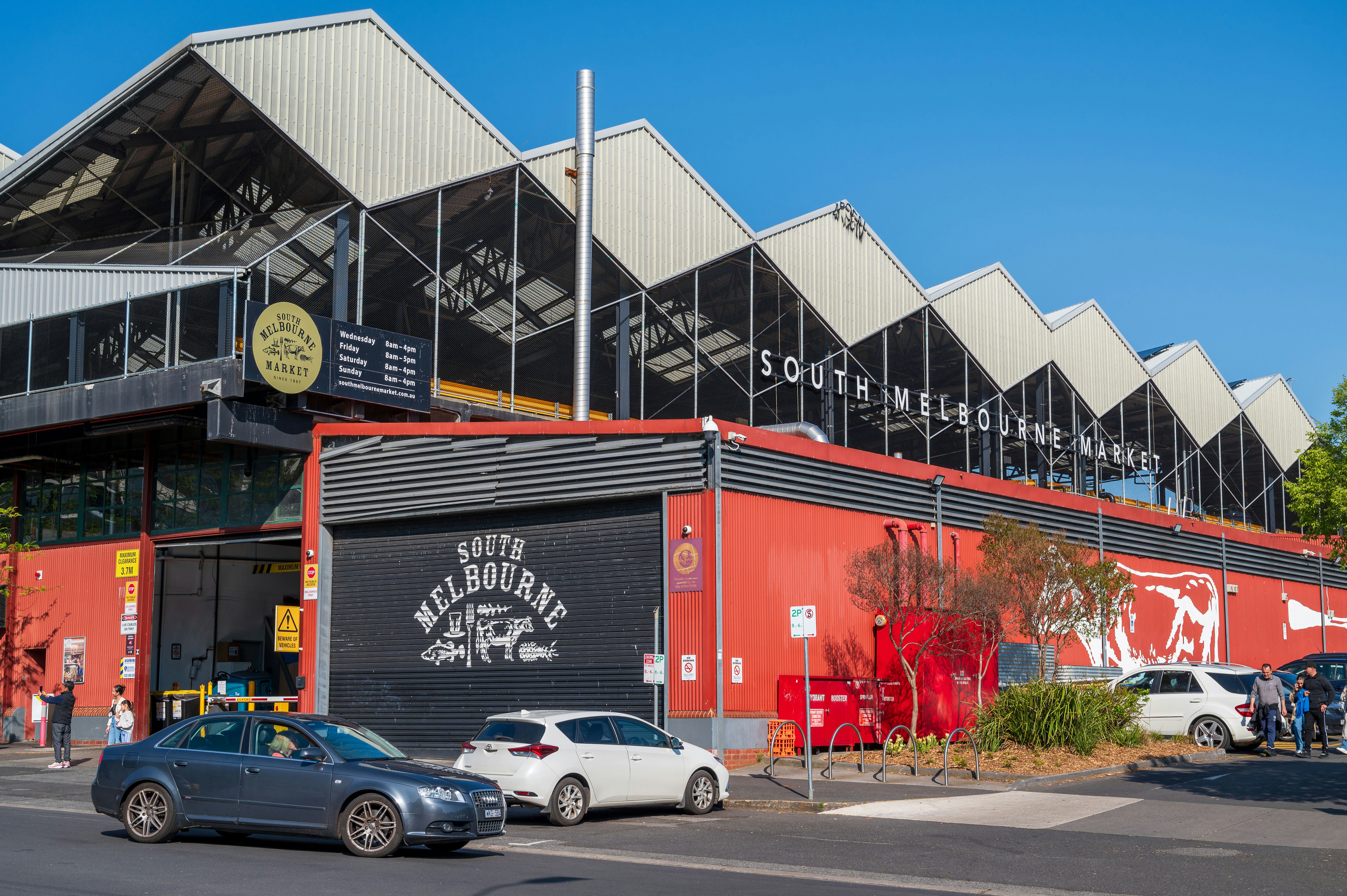 South Melbourne Market's distinctive sawtooth roof and vibrant red facade under a clear blue sky.