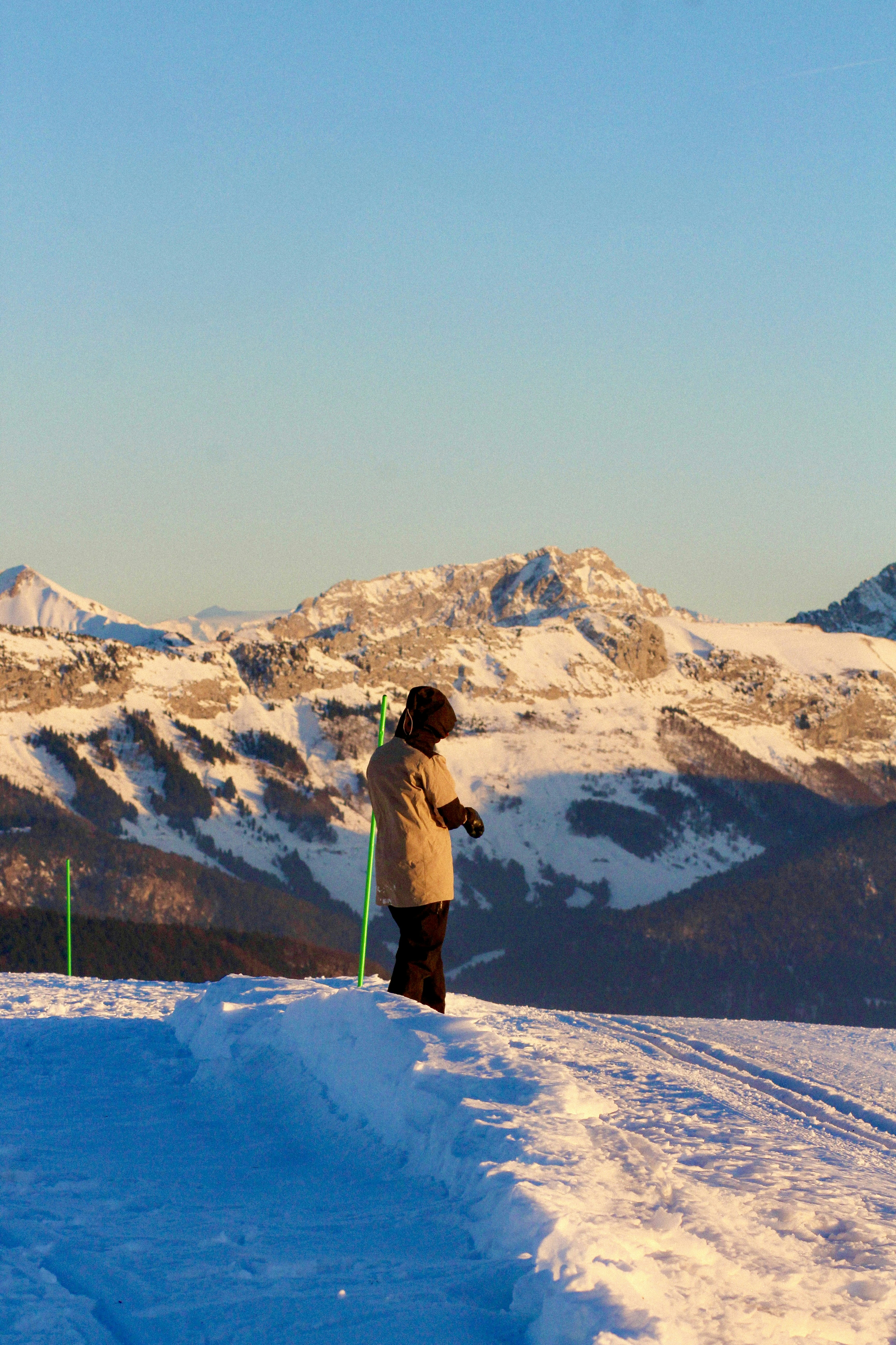 Person standing on a snow-covered mountain with distant peaks illuminated by the warm glow of sunset.