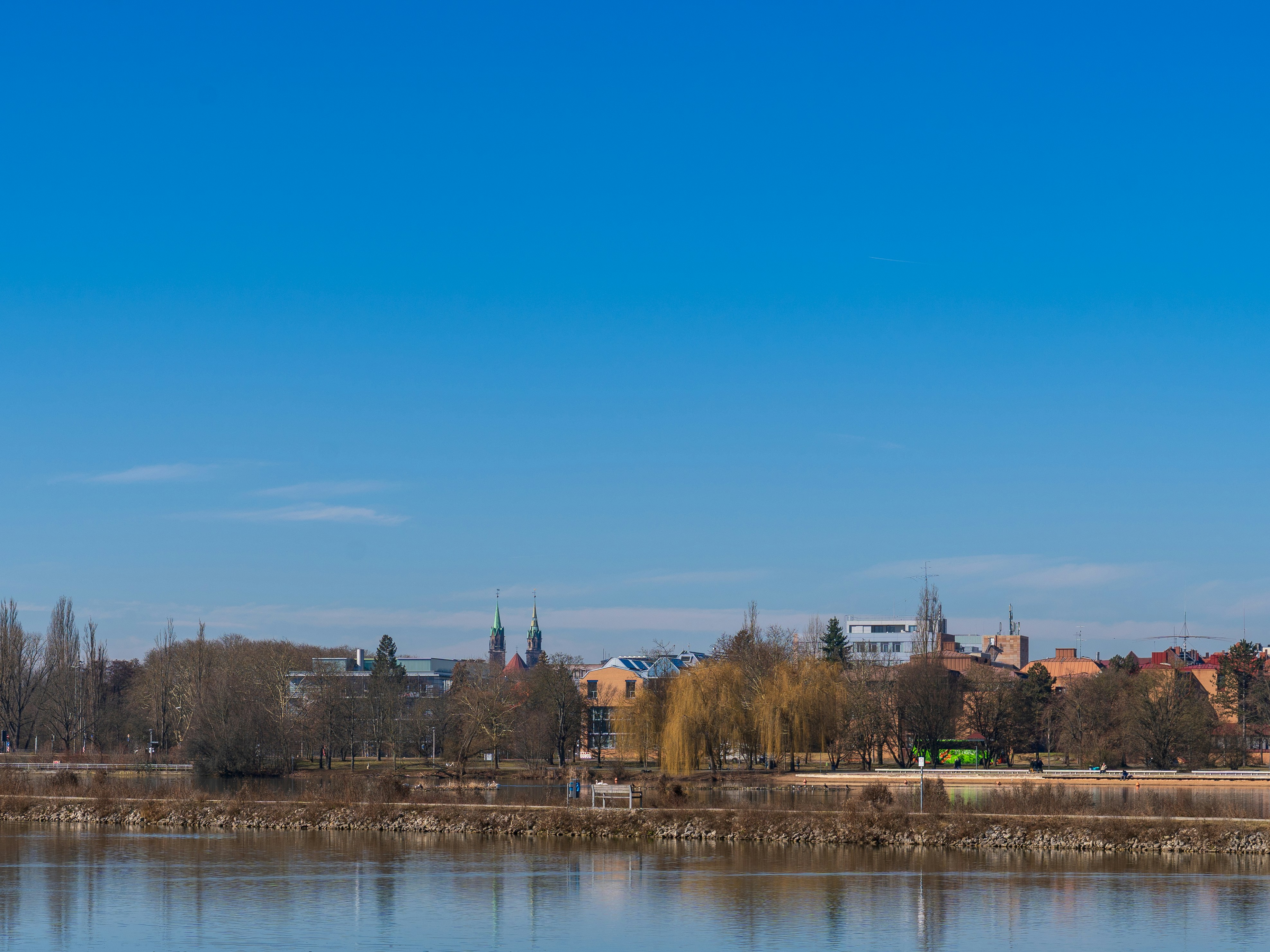 Serene lake reflecting urban architecture and bare trees beneath a vivid blue sky.