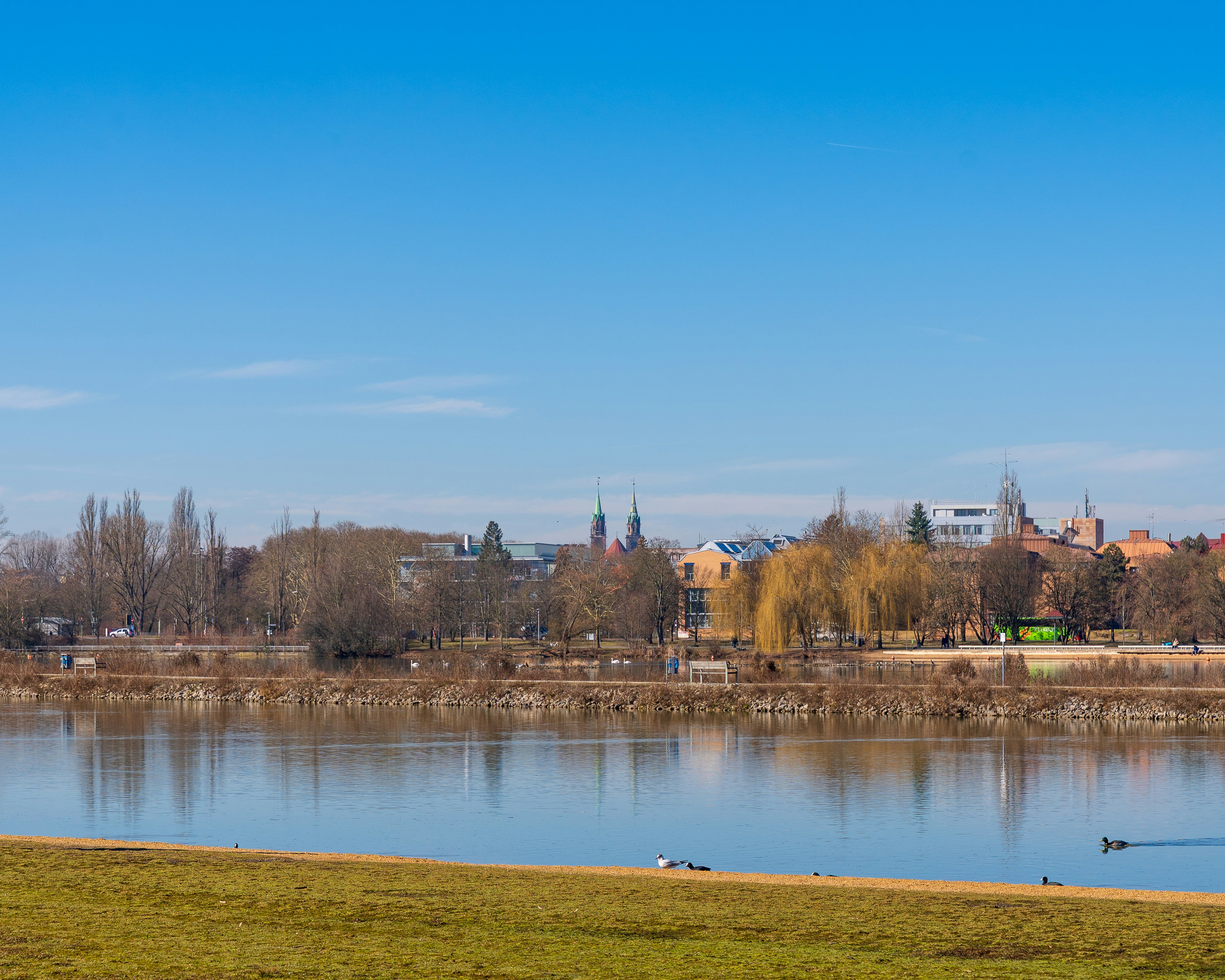 Calm lake reflecting a cityscape with trees and distant spires under a clear blue sky.