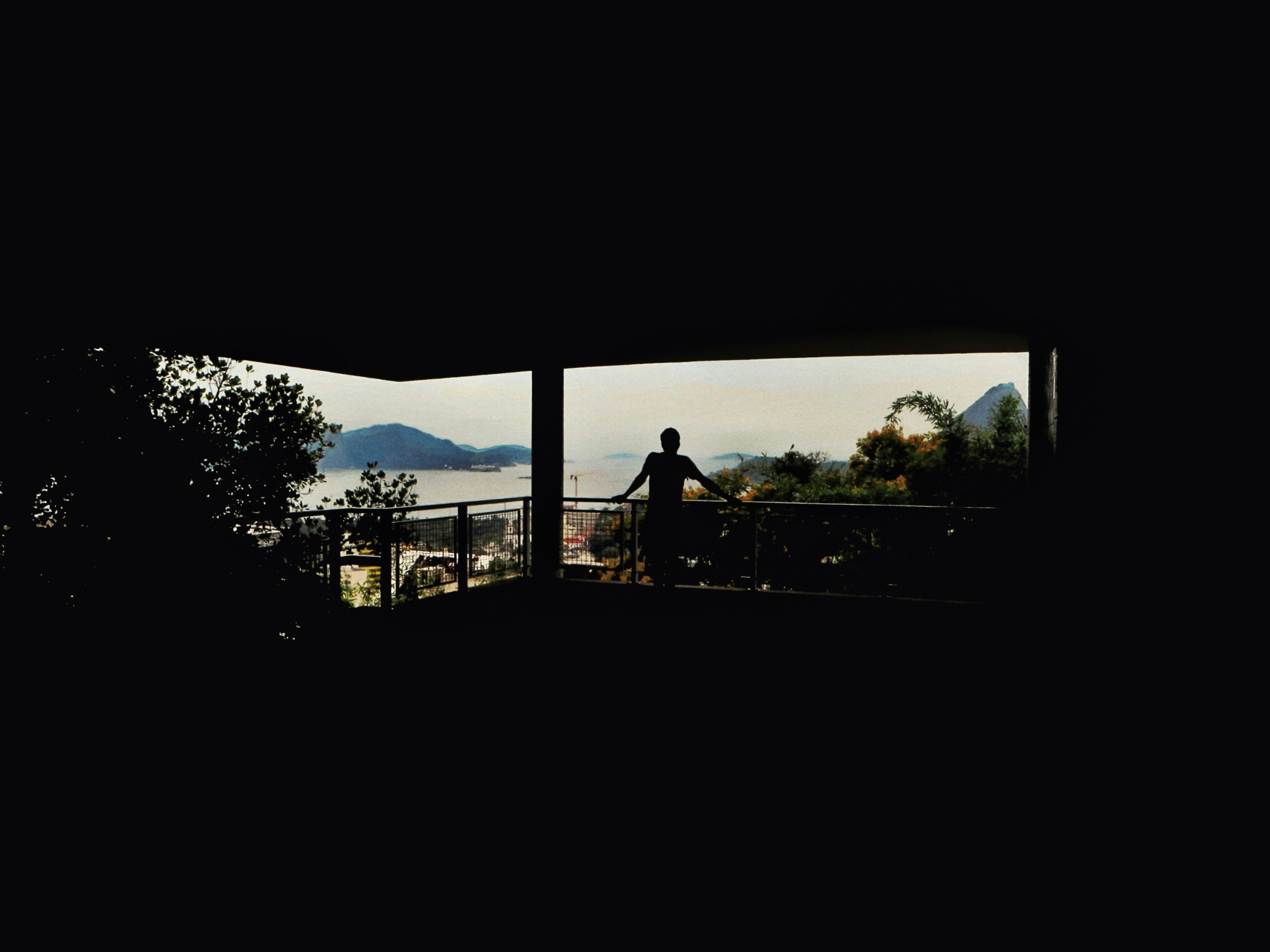 Silhouette of a person standing on a balcony overlooking distant hills and calm water.