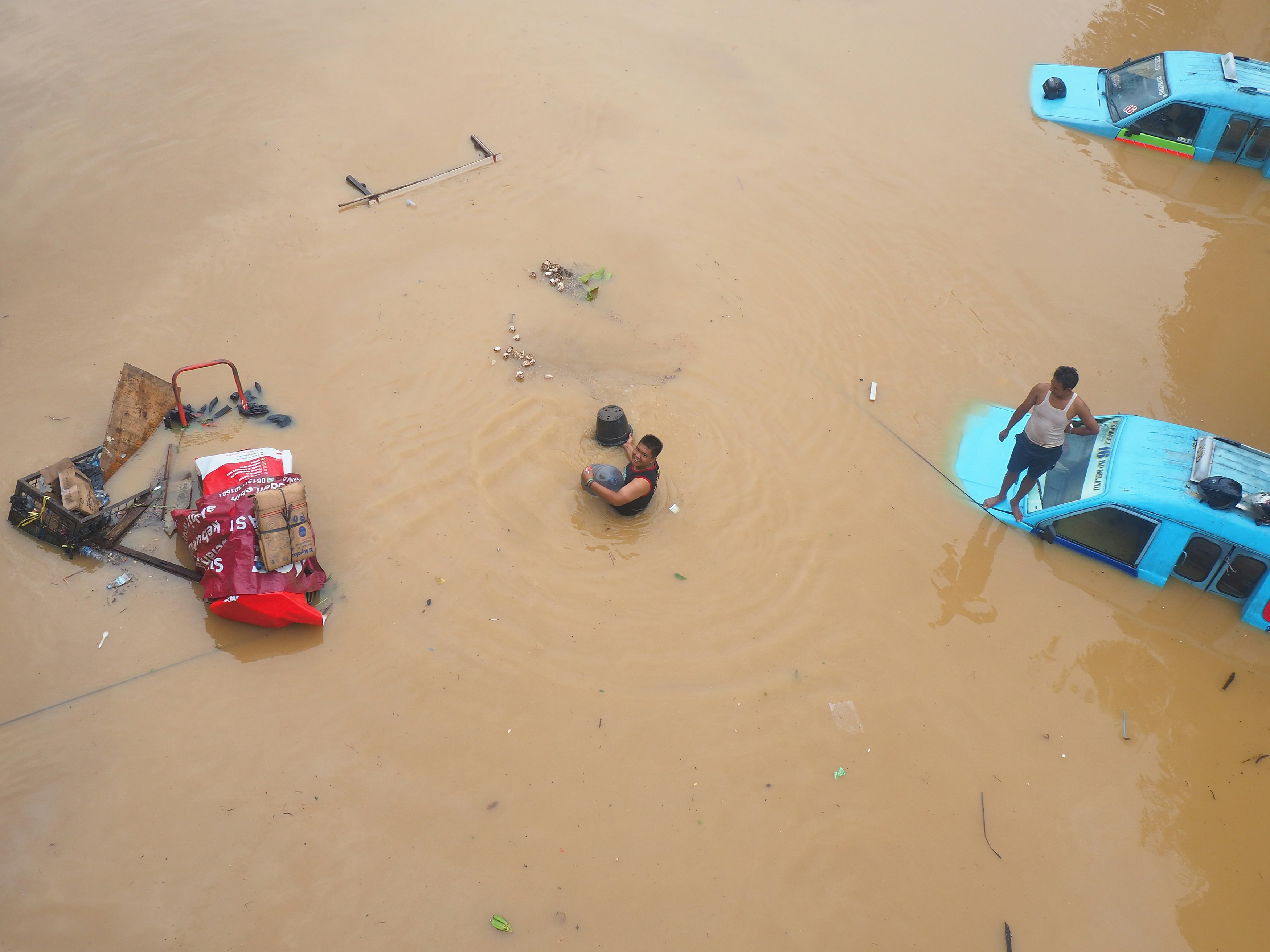 Two people navigate floodwaters in Jakarta, surrounded by submerged vehicles and debris.