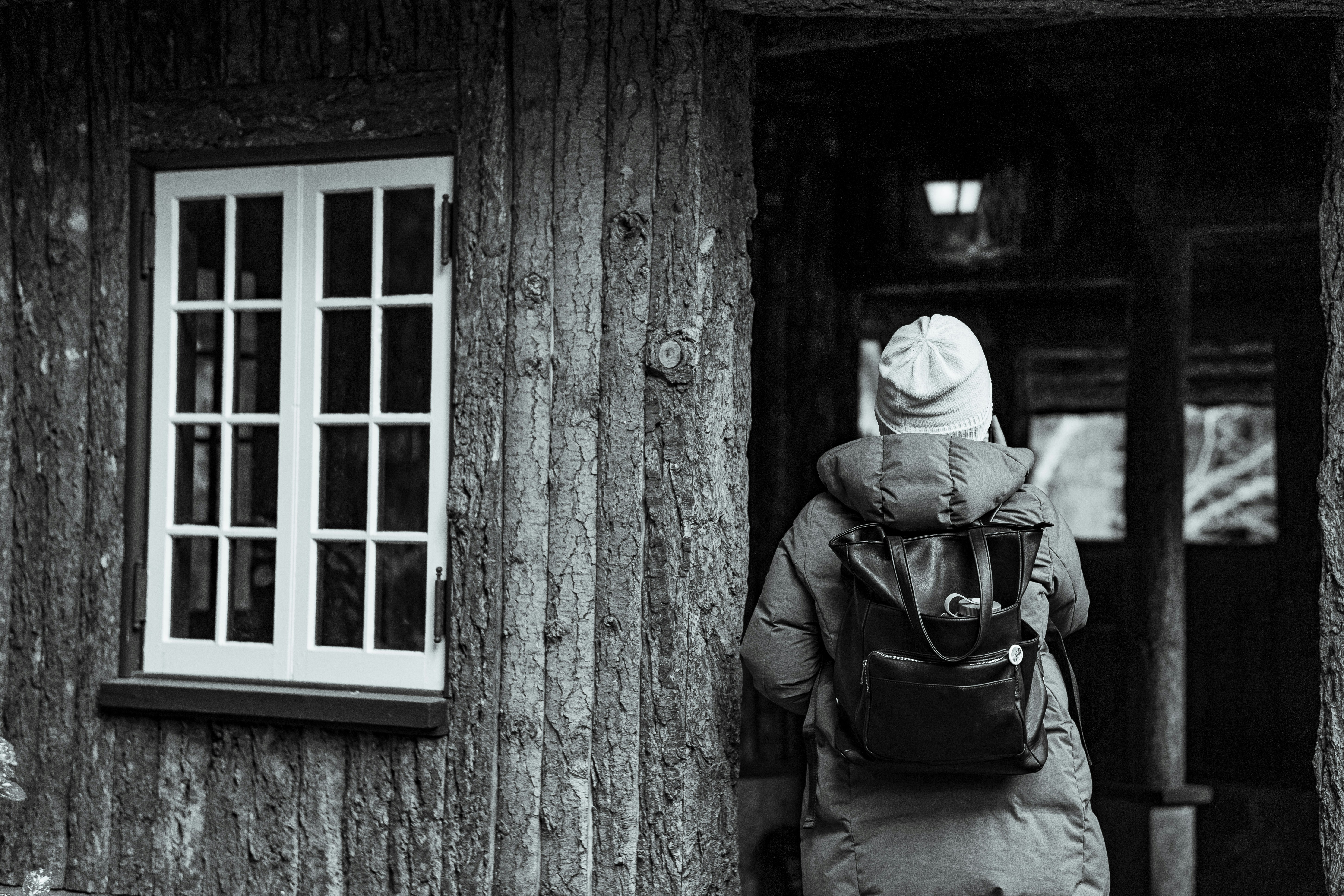 Person in winter attire facing a wooden cabin with a small window.