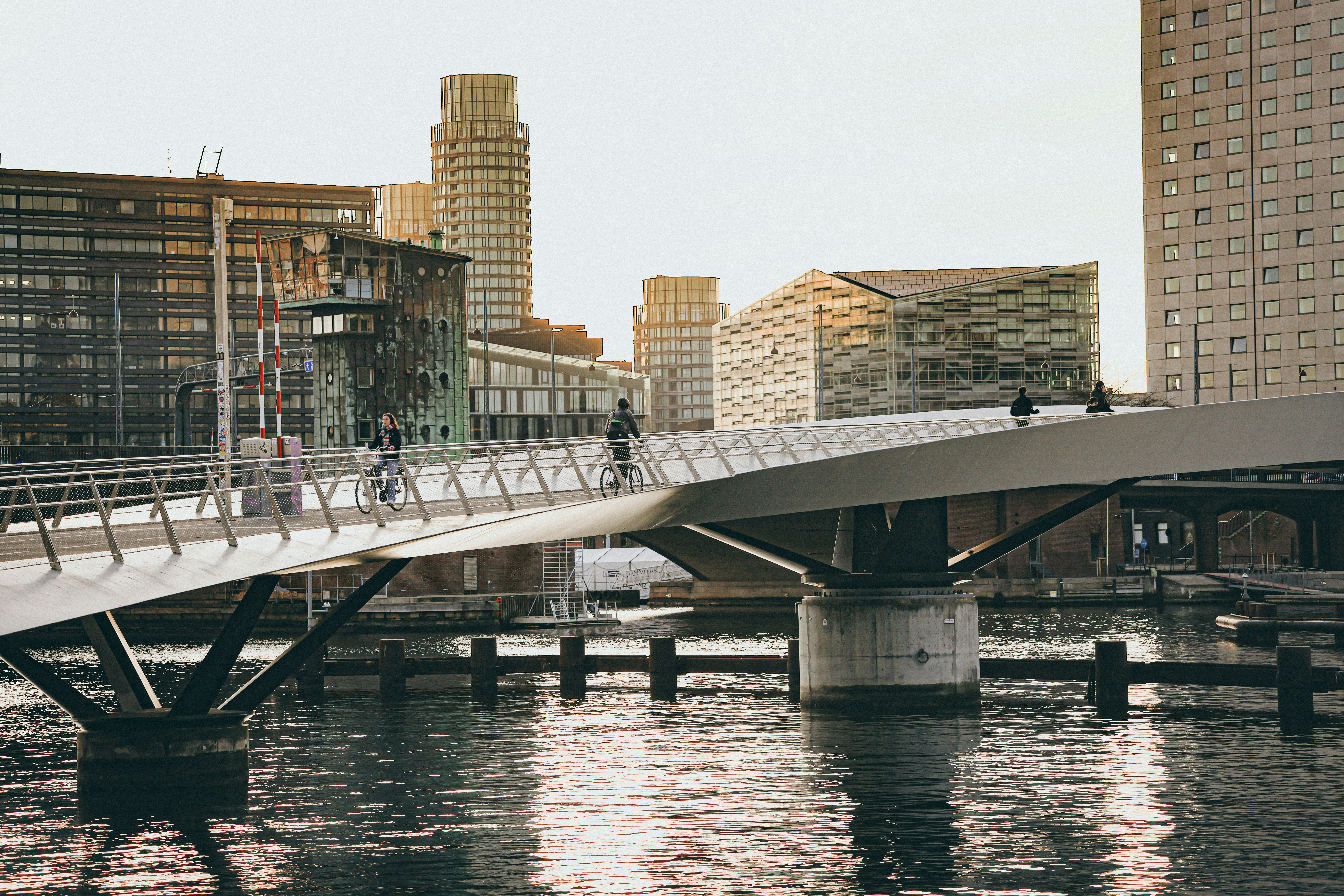 Cyclists traverse a sleek bridge against a backdrop of contemporary city architecture at sunset.
