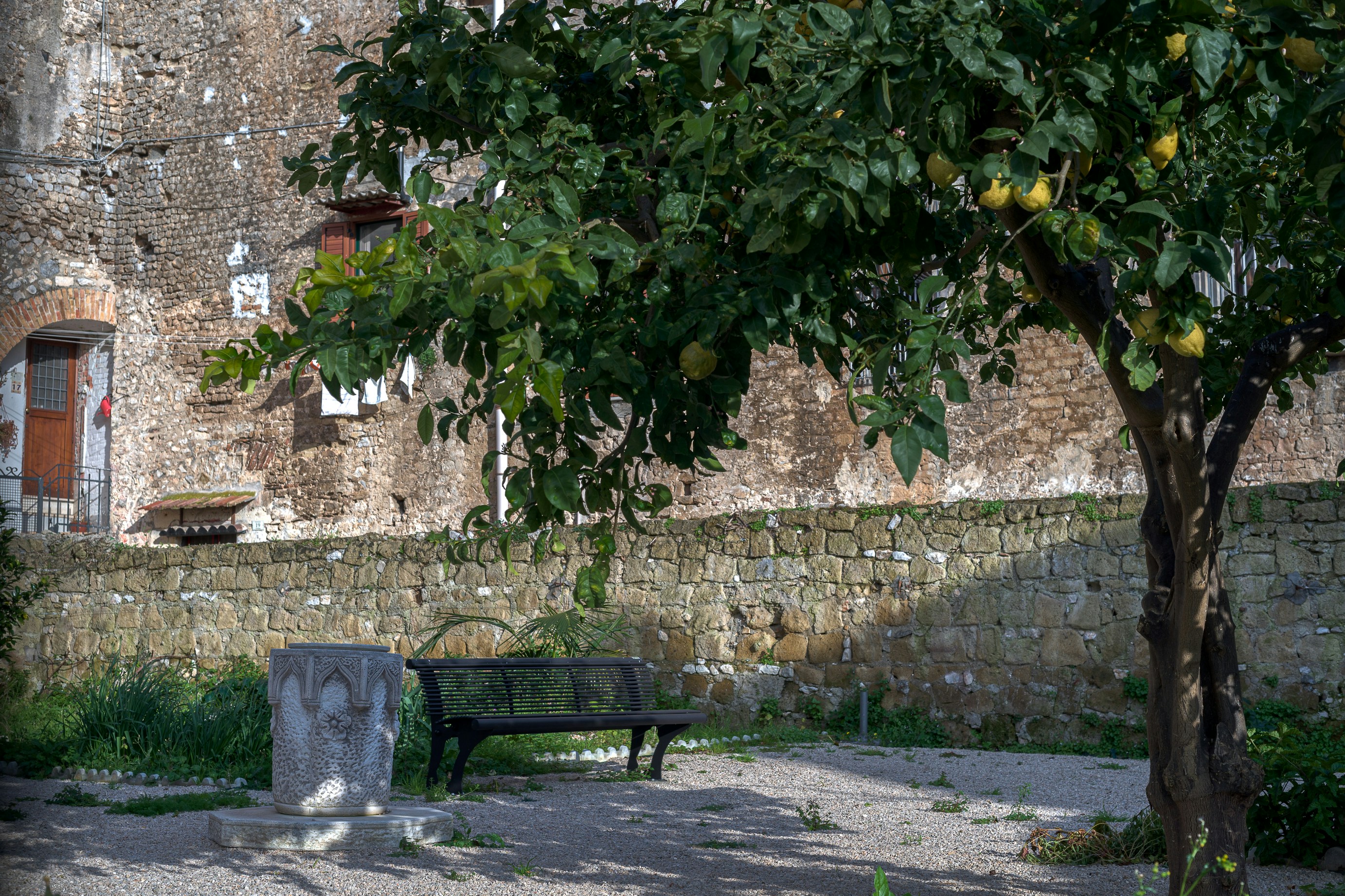 Courtyard in Terracina with a lemon tree casting shadows on a stone bench and ancient wall.