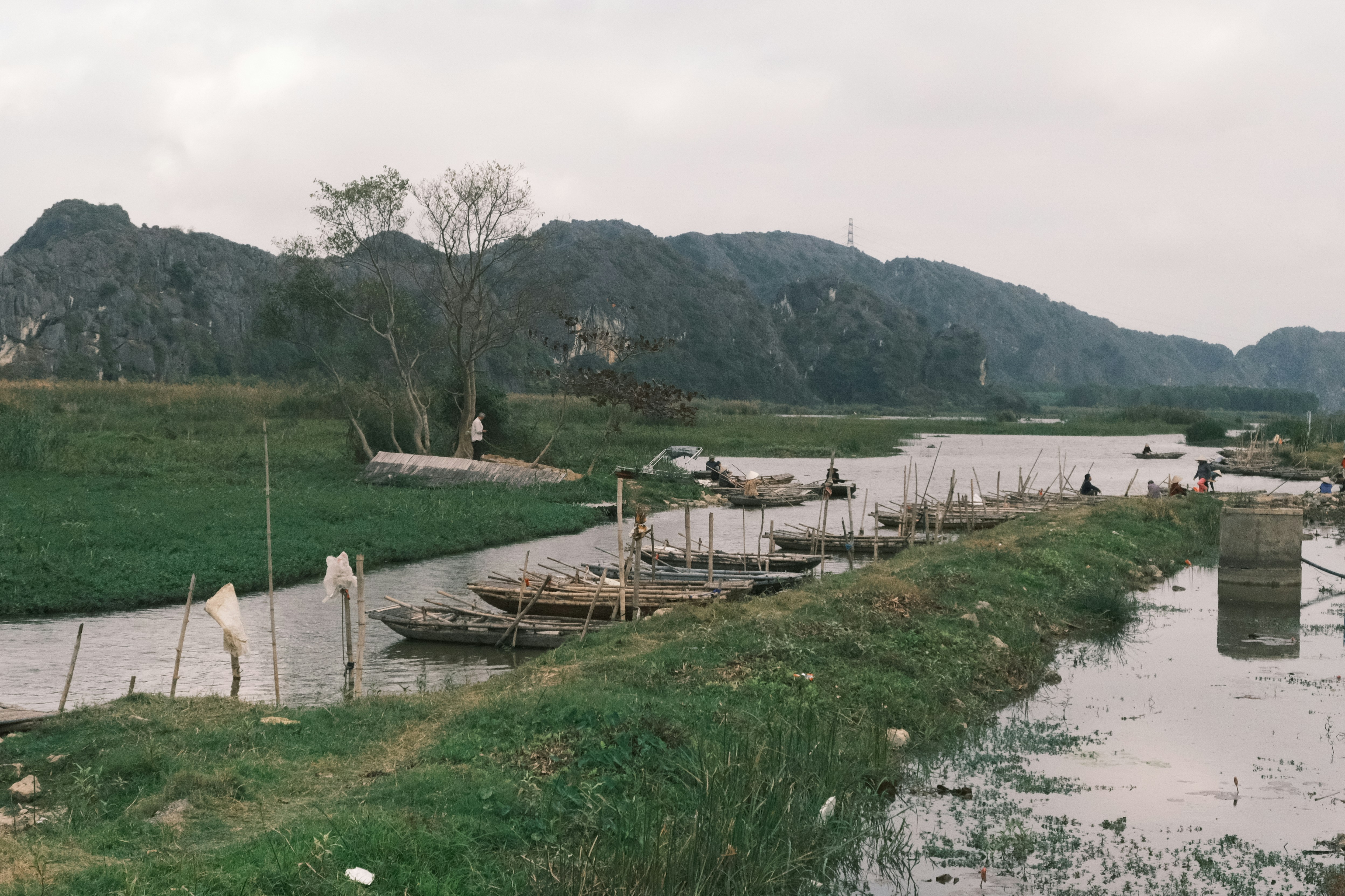 Wooden boats moored along a tranquil river with lush greenery and distant mountains under a cloudy sky.