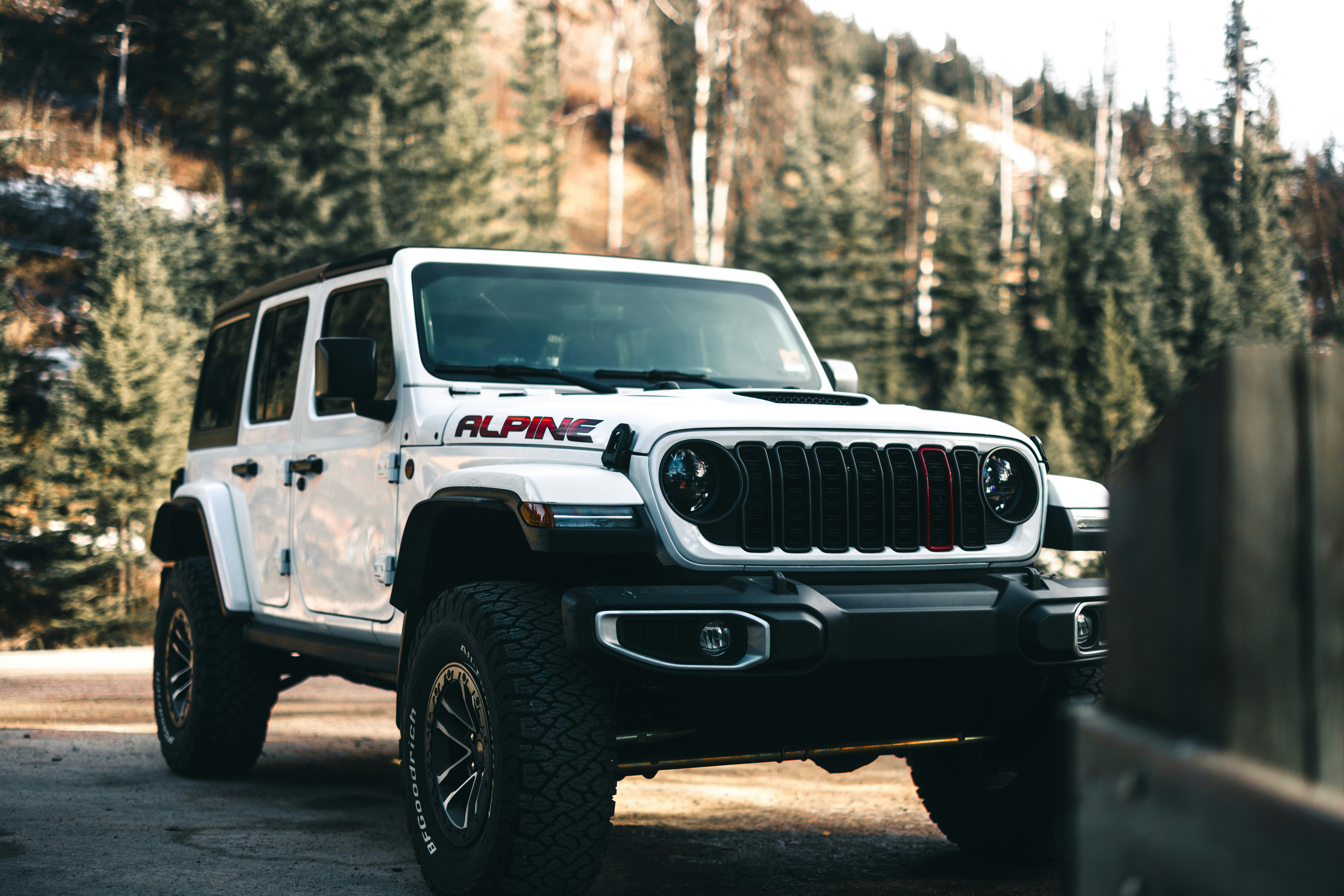 White off-road vehicle parked against a backdrop of tall evergreen trees in a mountainous setting.