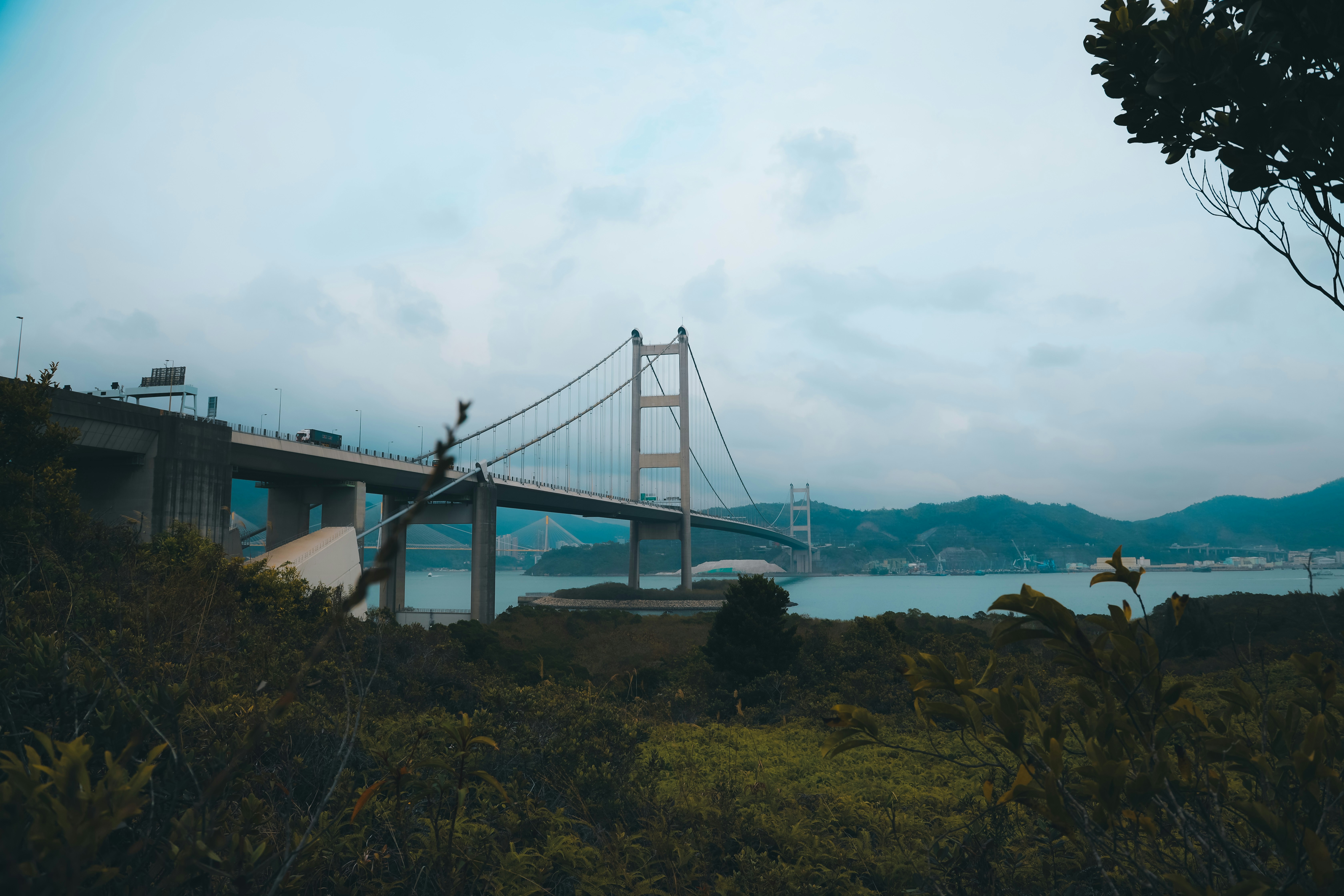 Suspension bridge stretches over a lush landscape with mountains in the background under a cloudy sky.