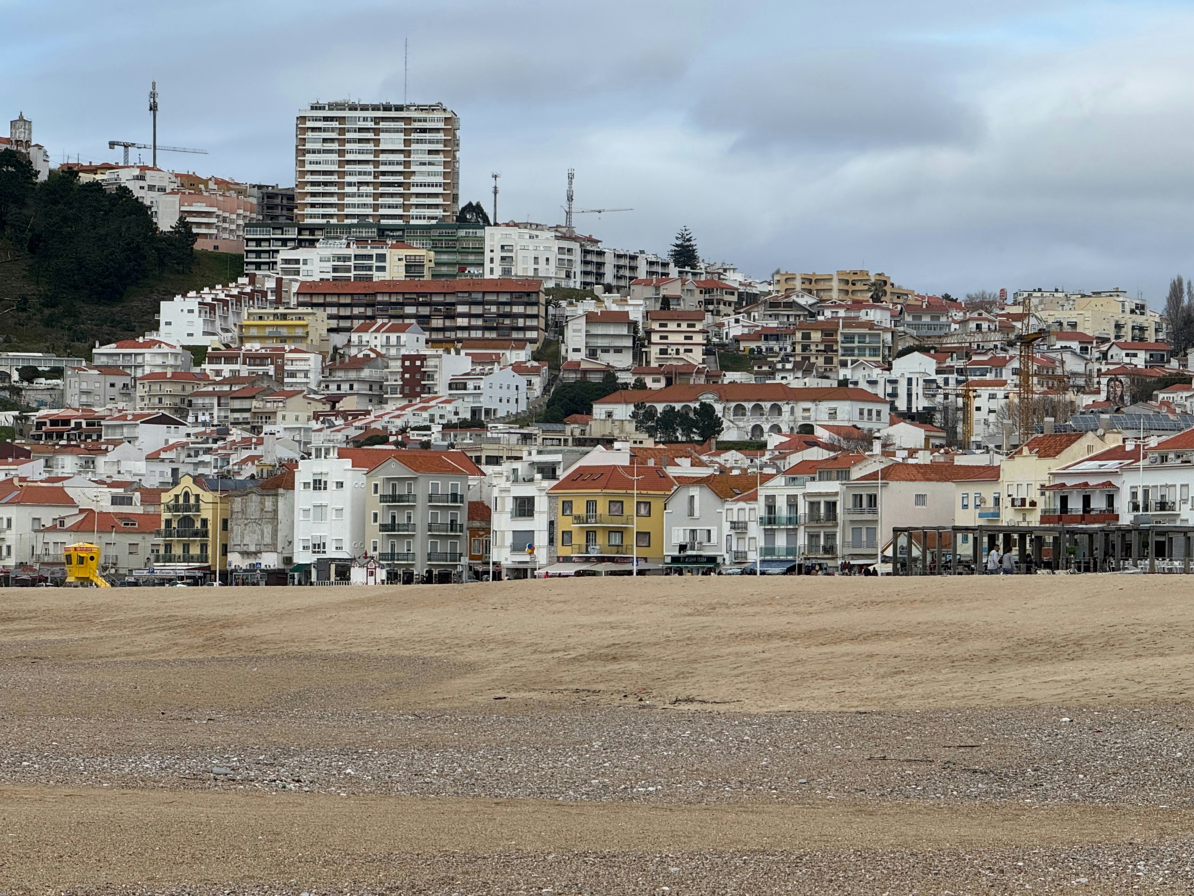 Clustered hillside buildings with red-tiled roofs overlooking a sandy beach under cloudy skies.