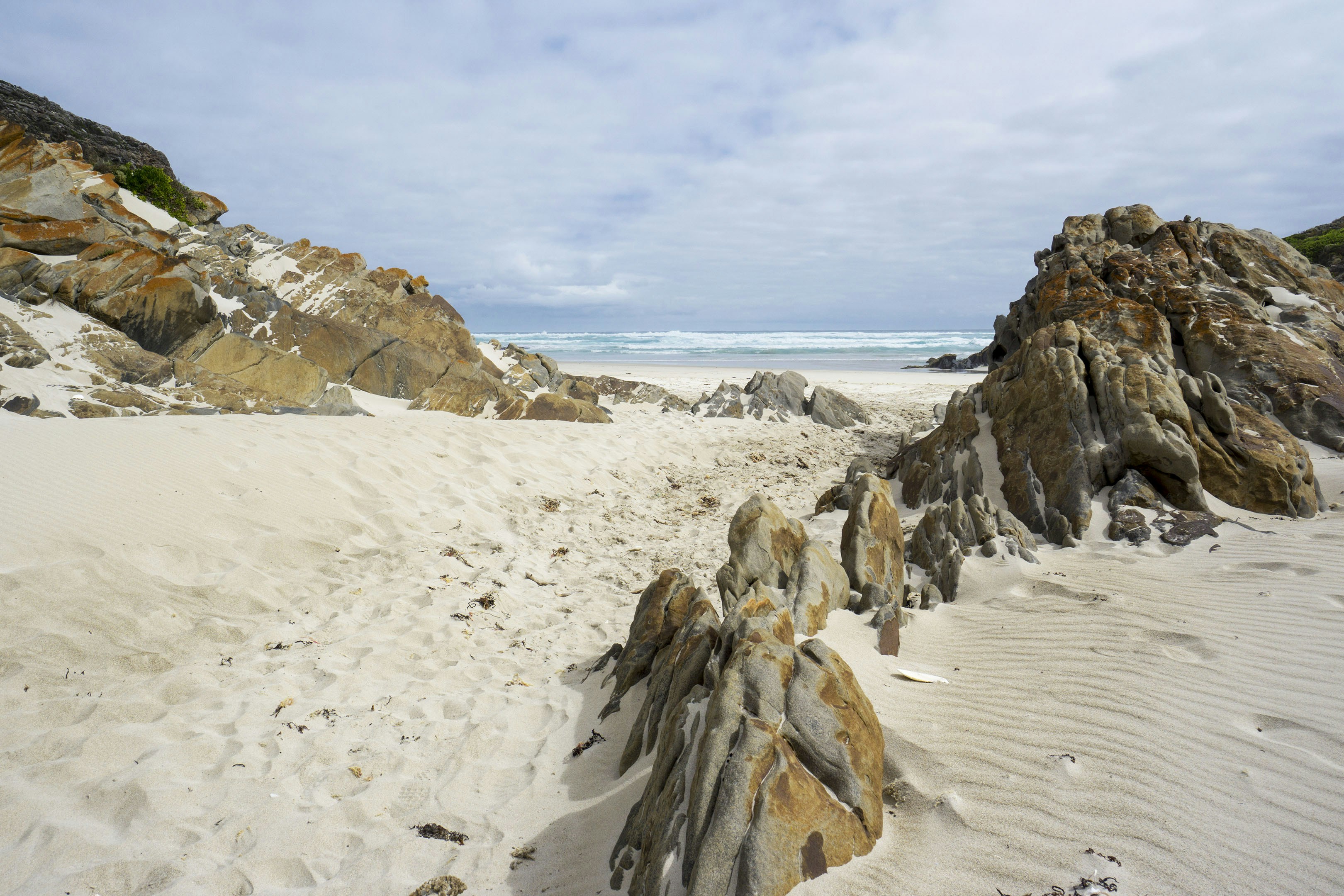 Sandy beach path flanked by rugged rock formations leading to ocean waves under a cloudy sky.